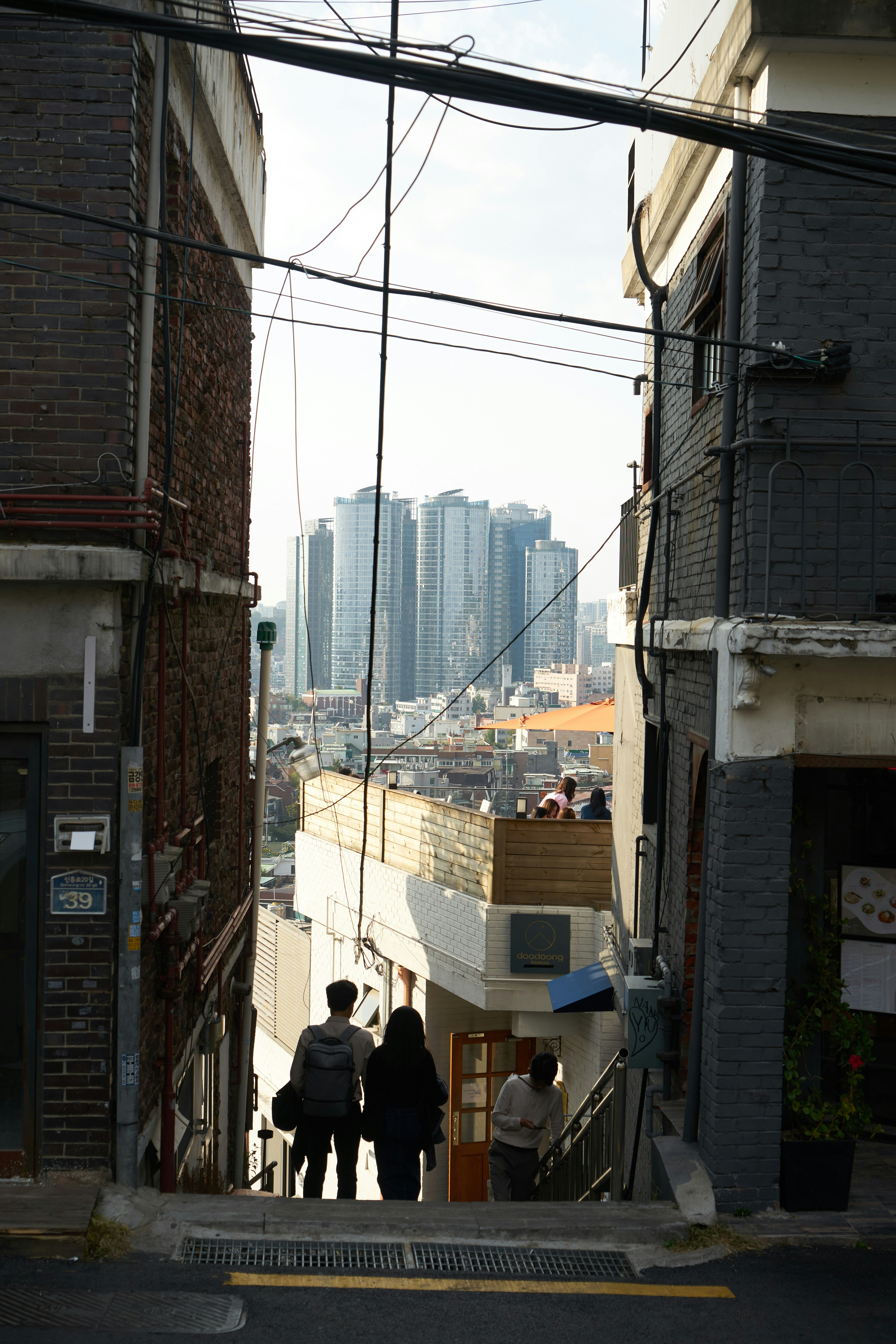 Two figures walking down a narrow street framed by urban buildings, leading towards a modern city skyline in the background.