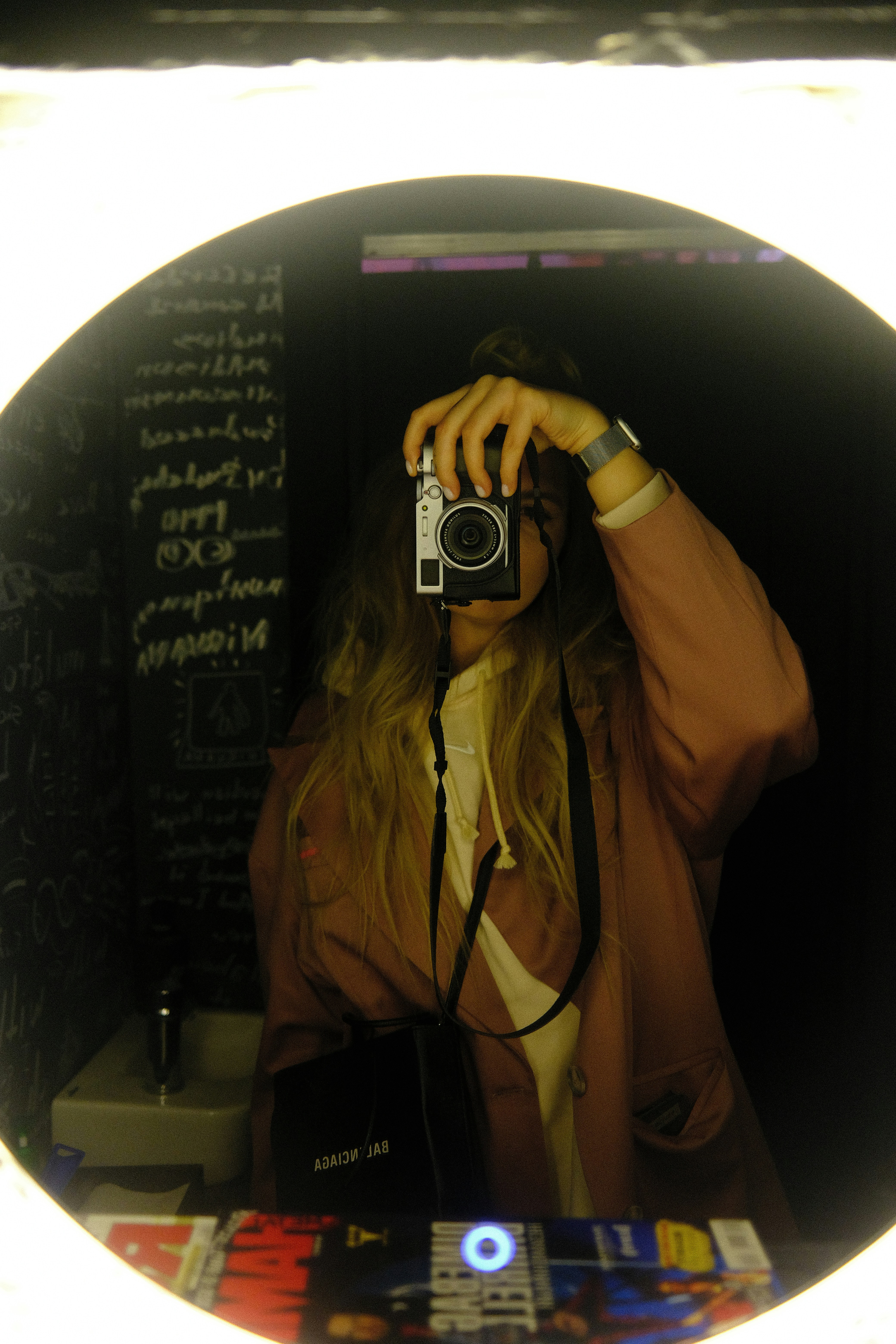 A person with long hair holding a camera, reflected in a circular mirror surrounded by dark walls filled with writings.