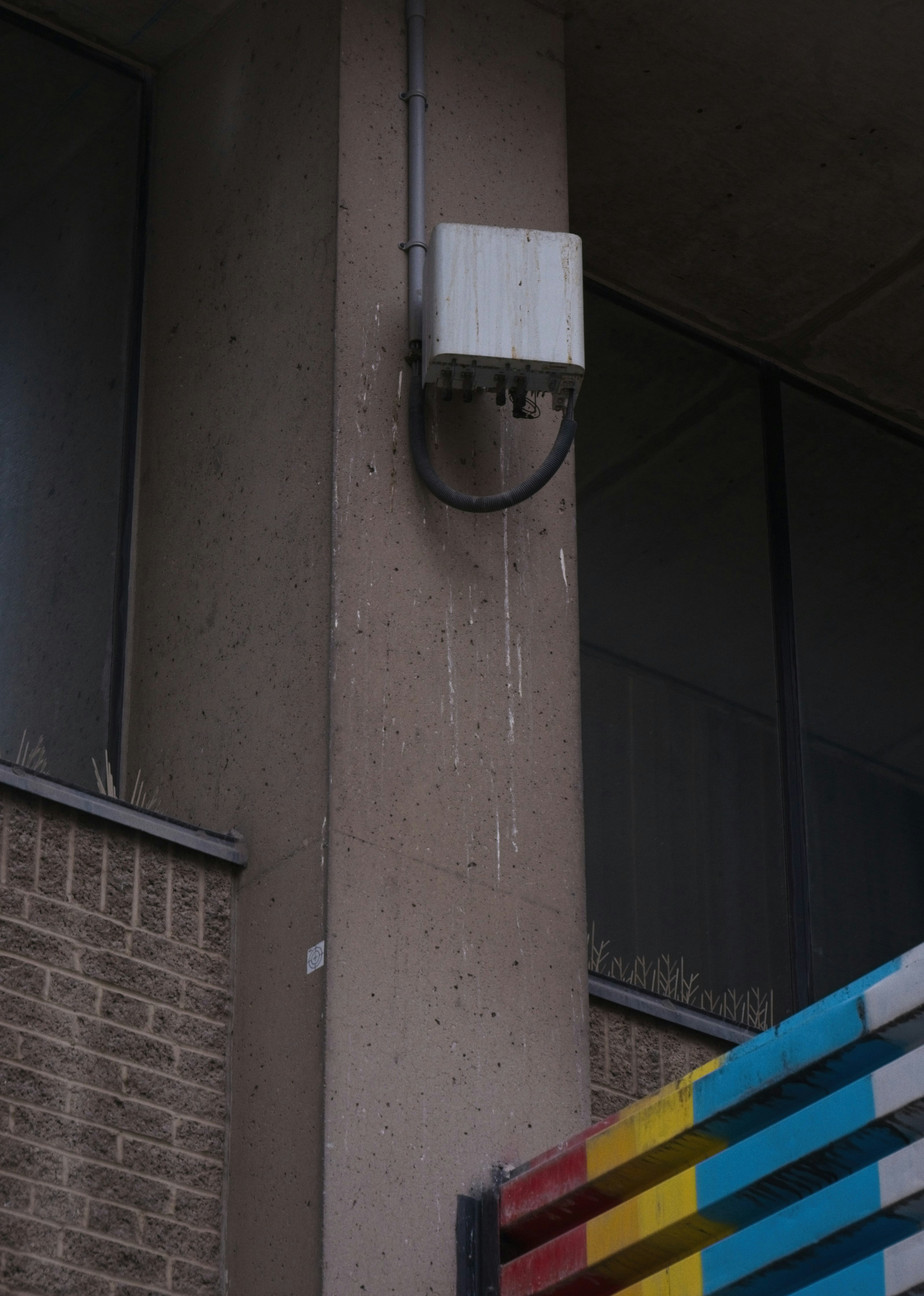 A textured concrete wall features a mounted device alongside colorful horizontal stripes, highlighting urban architectural elements.