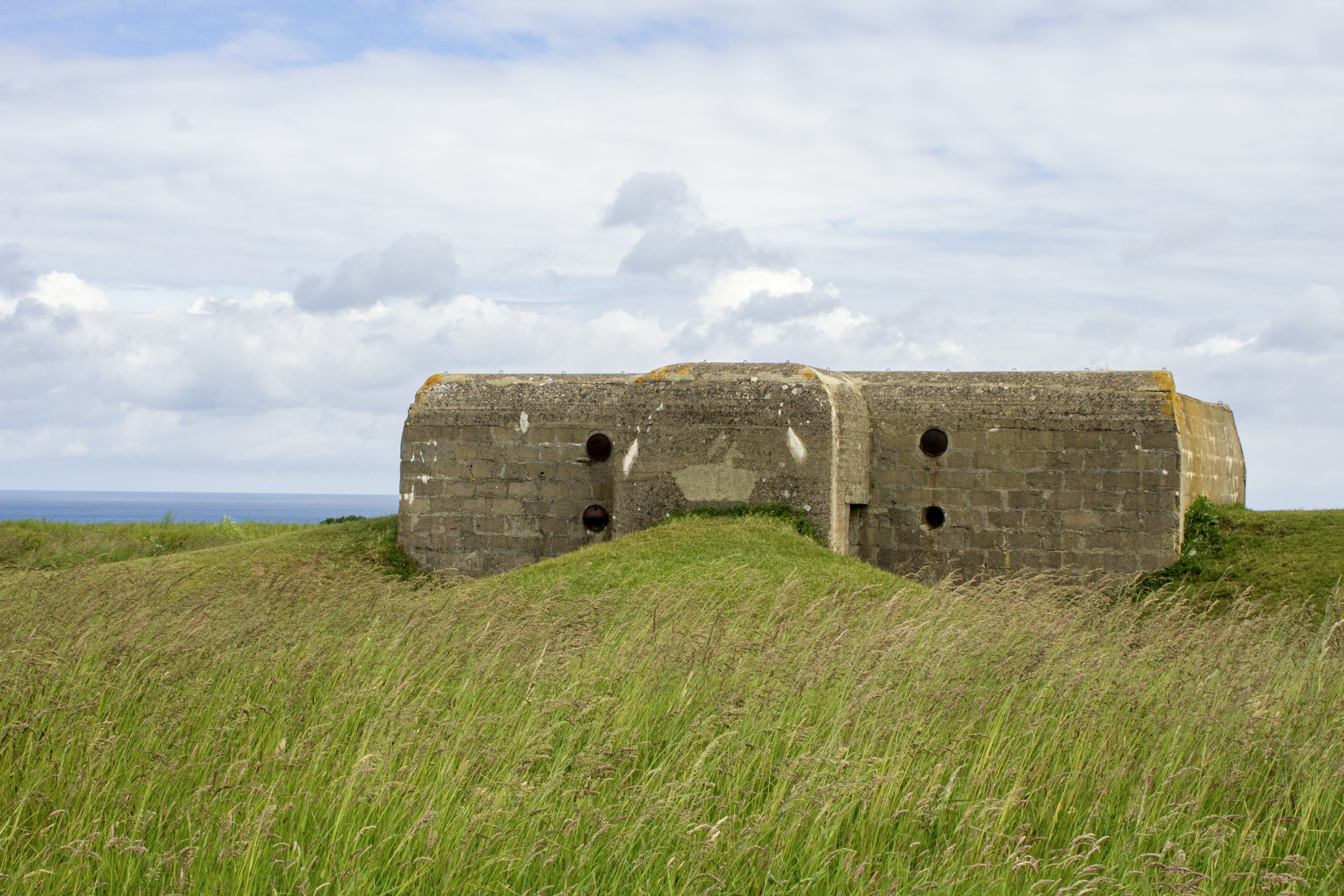 Weathered military bunker nestled in tall grass, overlooking the sea under a cloudy sky.