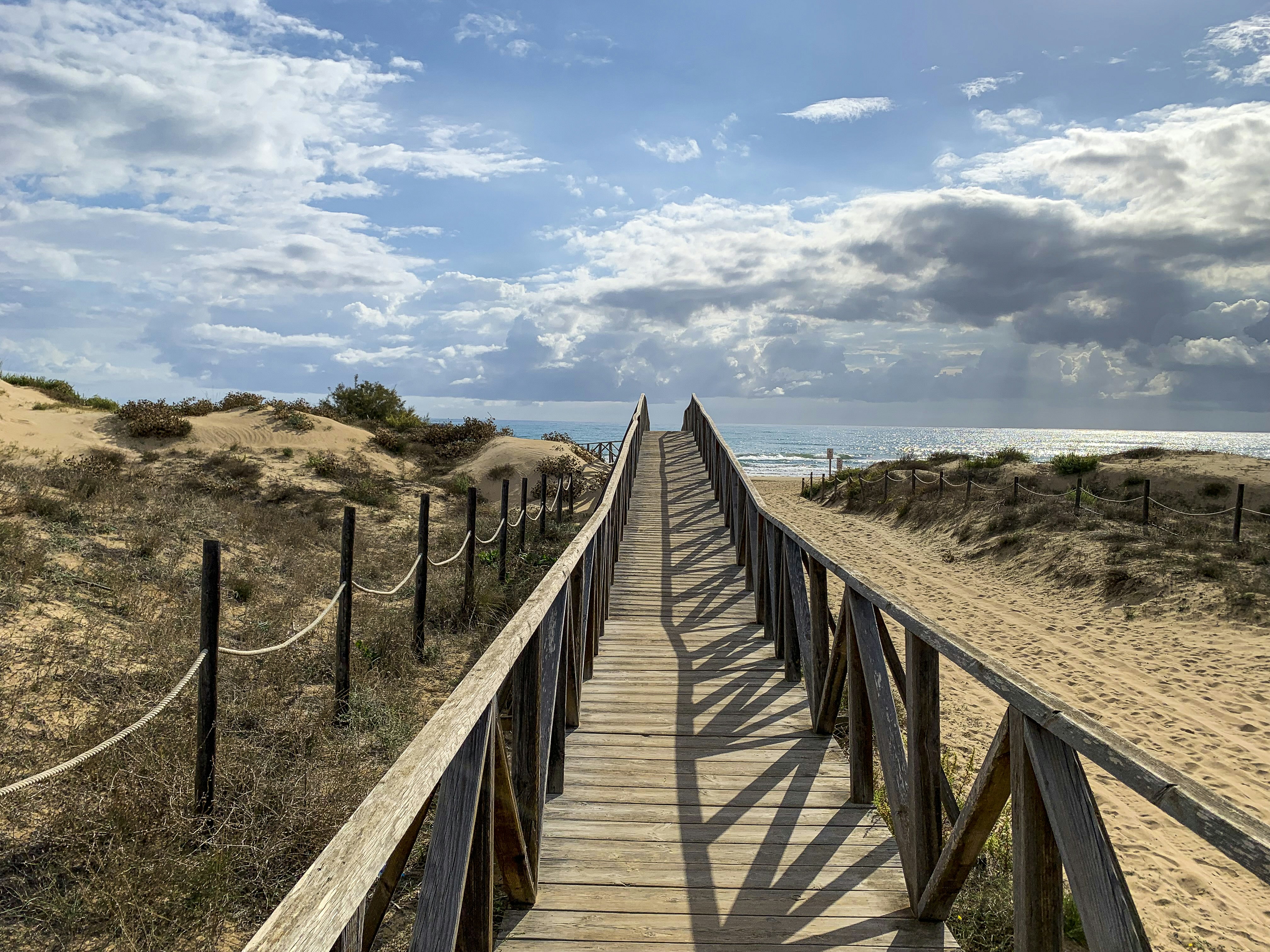 Wooden boardwalk leading through sandy dunes towards the ocean under a partly cloudy sky.
