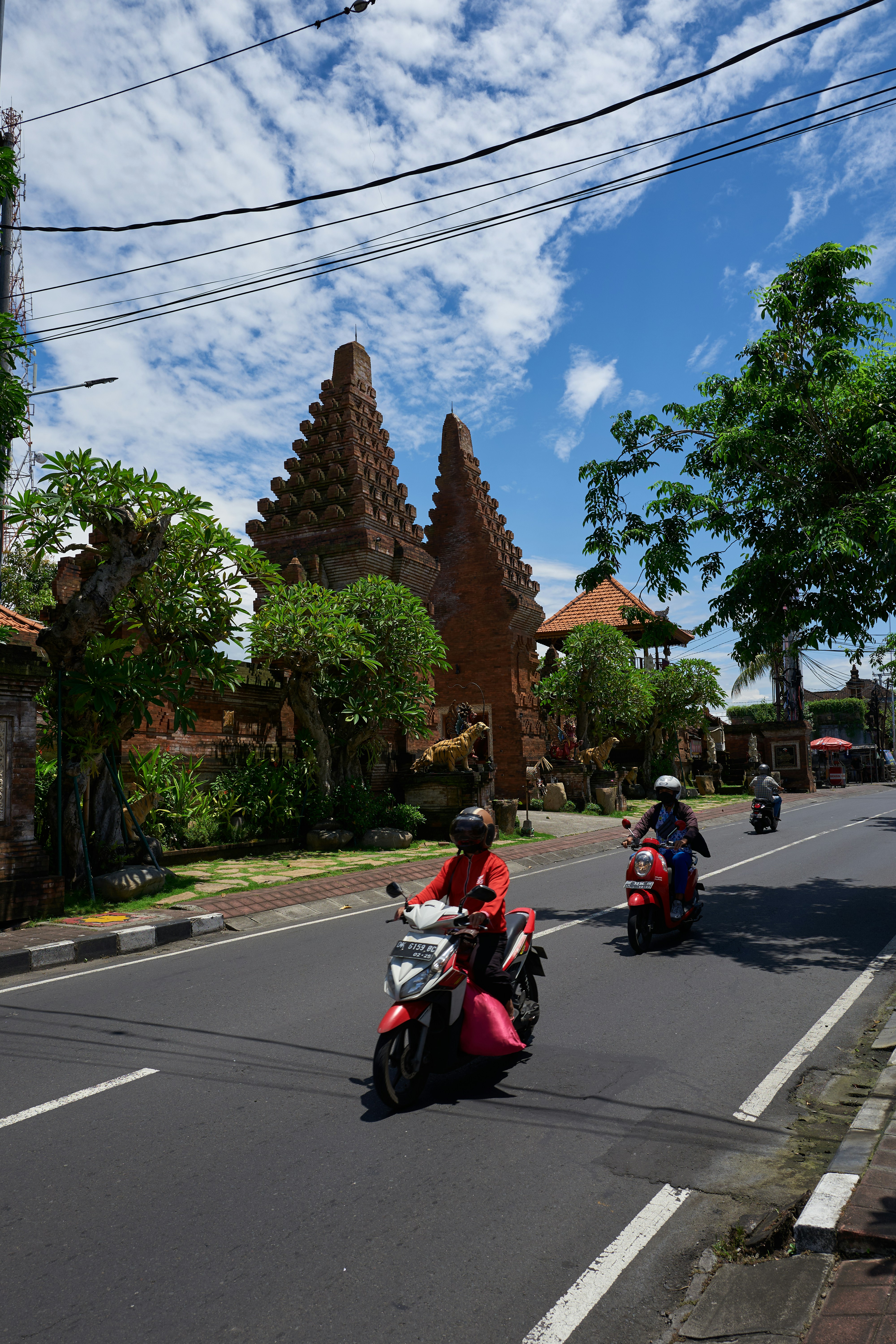 Motorcyclists navigate a vibrant street framed by traditional Balinese architecture and lush greenery.