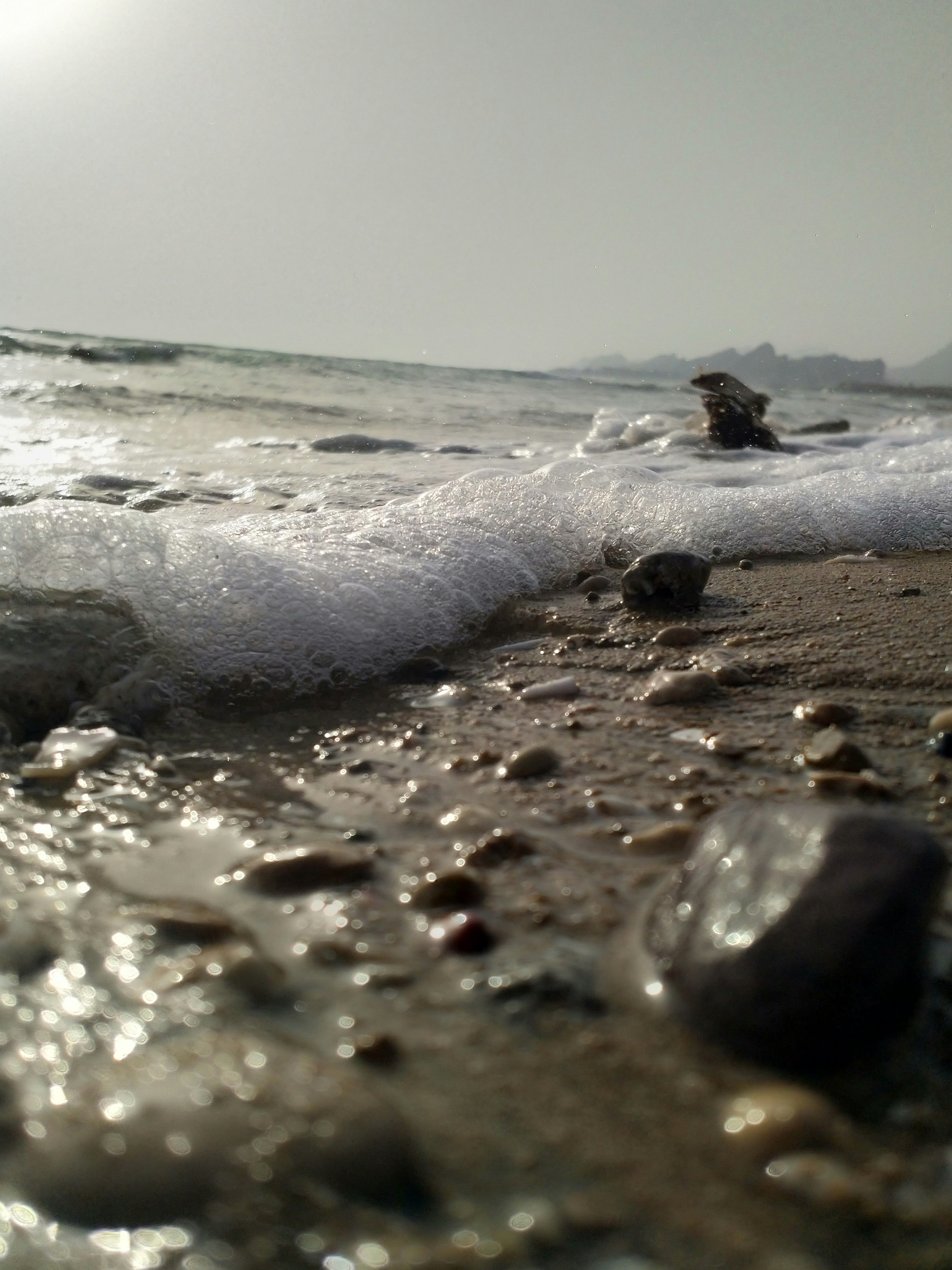Low-angle close-up of a pebbled shoreline as foamy surf rolls over wet sand toward the foreground. The scene highlights textures of stones, dark wet sand, and a distant, calm horizon under a pale sky.