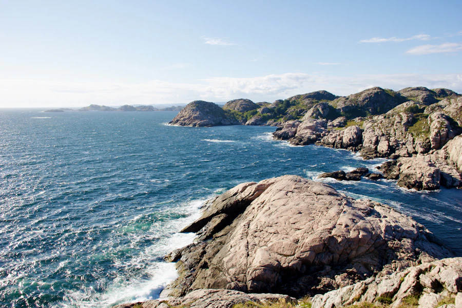 Sunlit rocky coastline with blue ocean waves curling around rugged cliffs and small offshore islands under a clear sky.