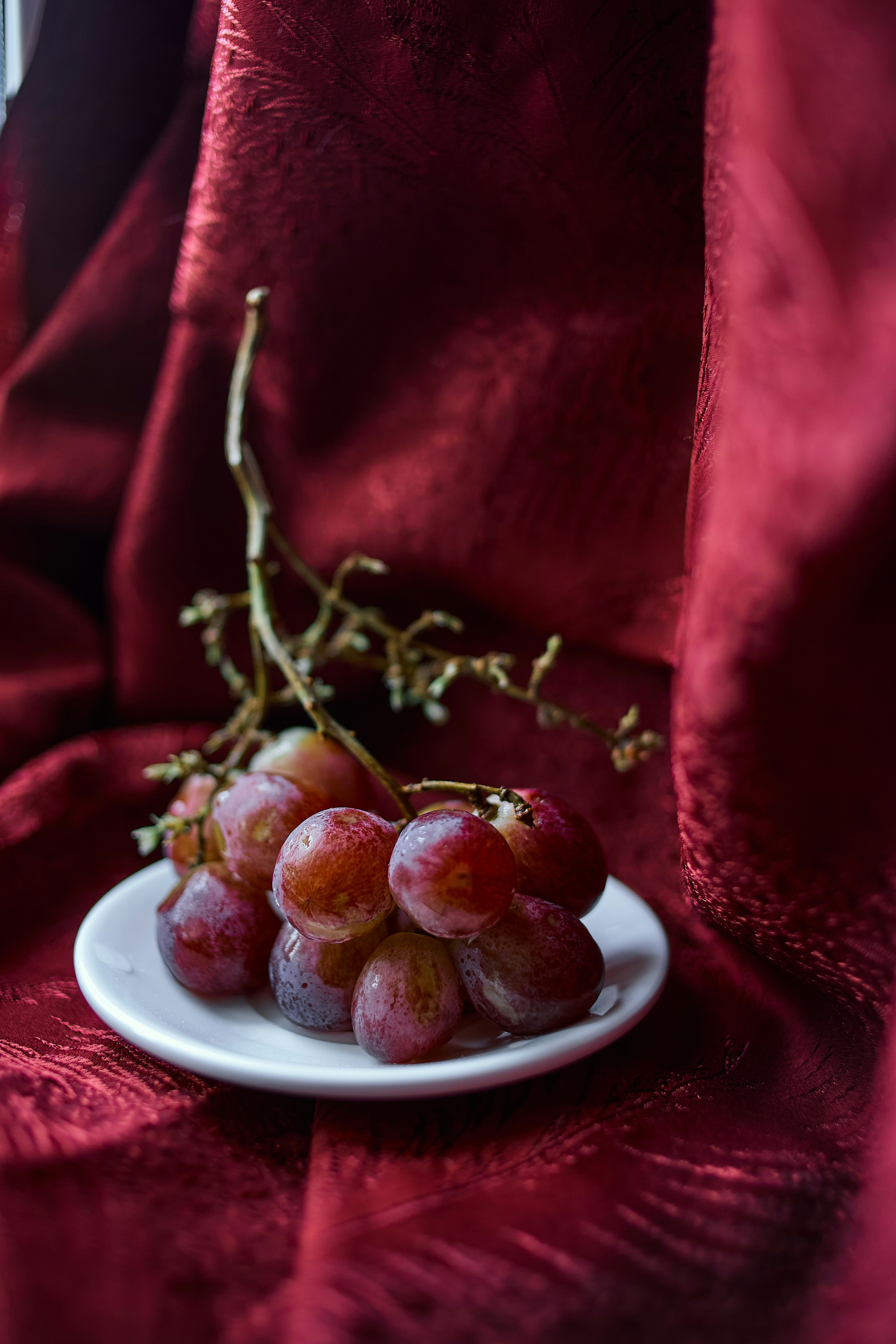 A cluster of ripe grapes on a small white plate set against luxurious scarlet fabric.