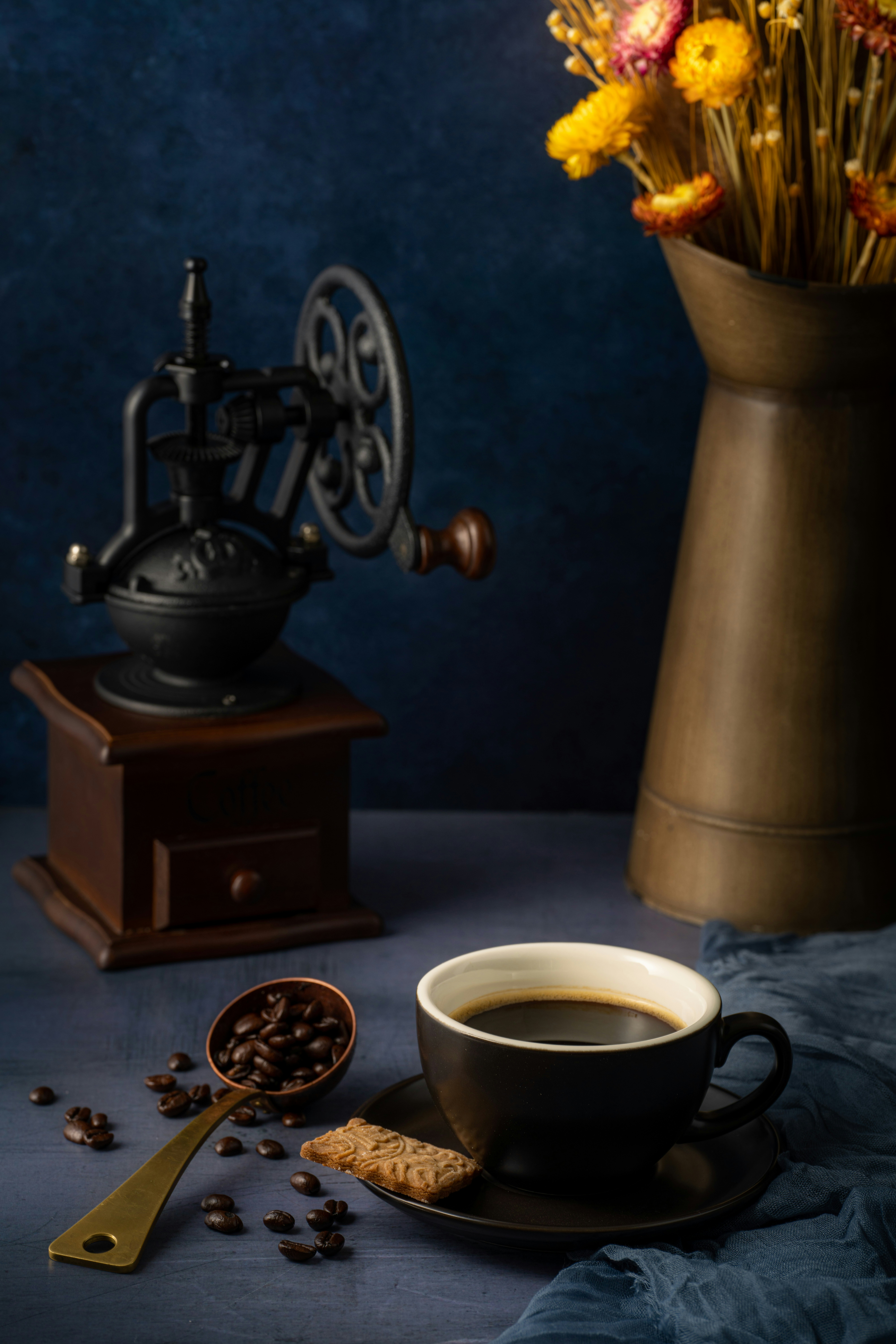 A dark coffee cup sits on a saucer beside a gold spoon filled with coffee beans, complemented by a vintage coffee grinder and a vase of dried flowers. 