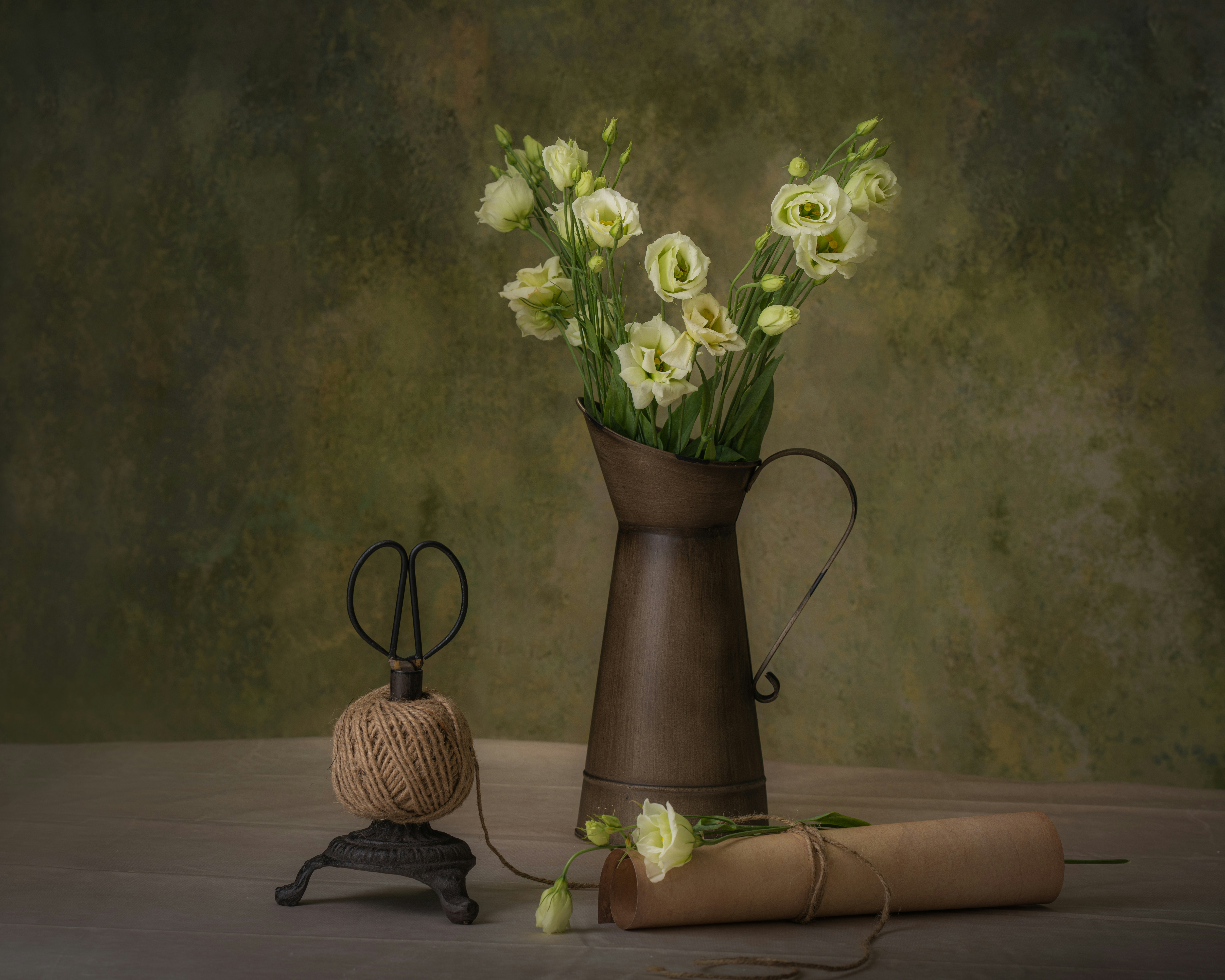 Delicate white flowers arranged in a vintage pitcher, accompanied by twine and a rolled parchment, set against a textured backdrop.