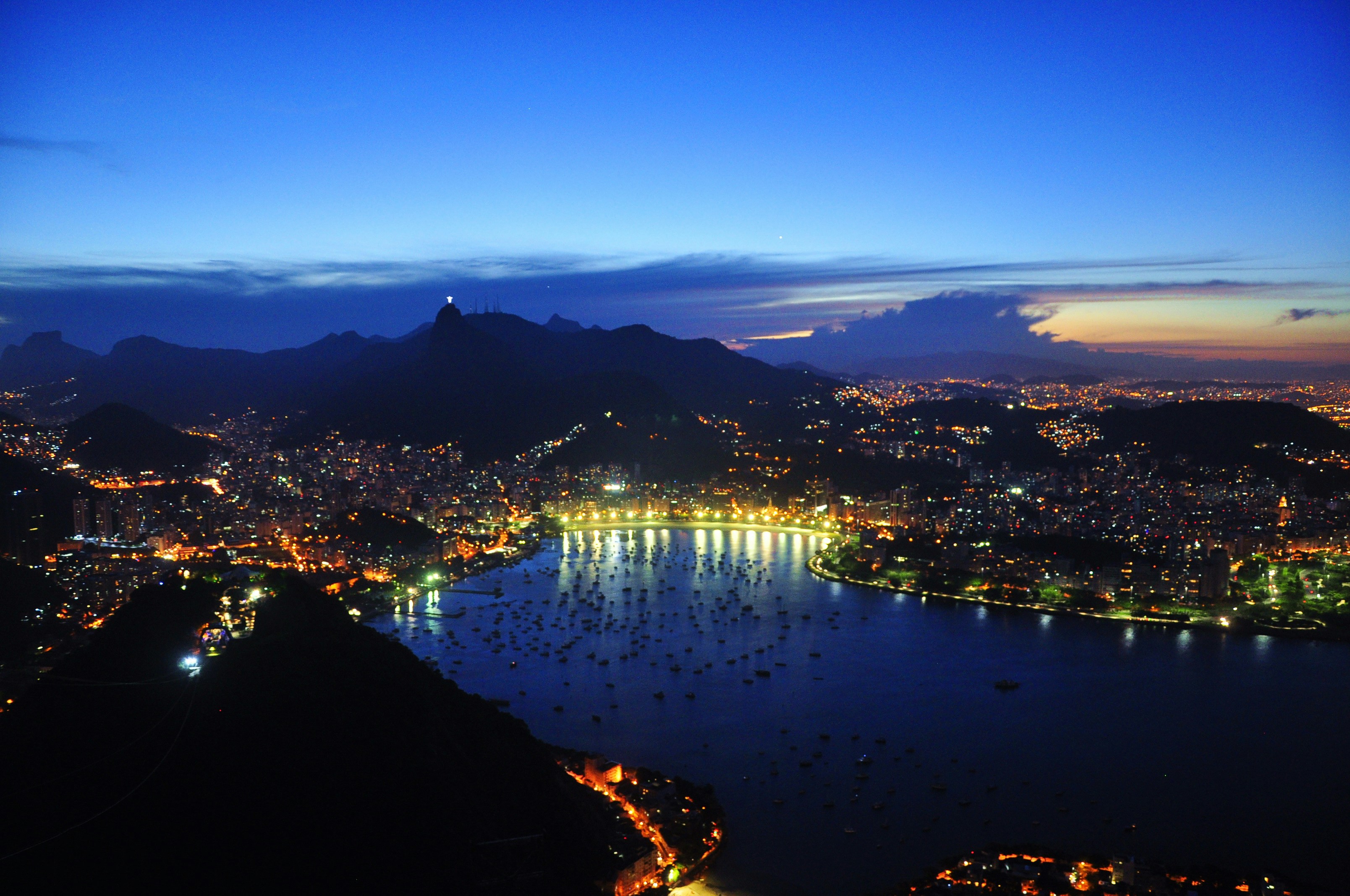 A panoramic view of Guanabara Bay at twilight, showcasing the city lights reflecting on the water's surface with mountains in the background.