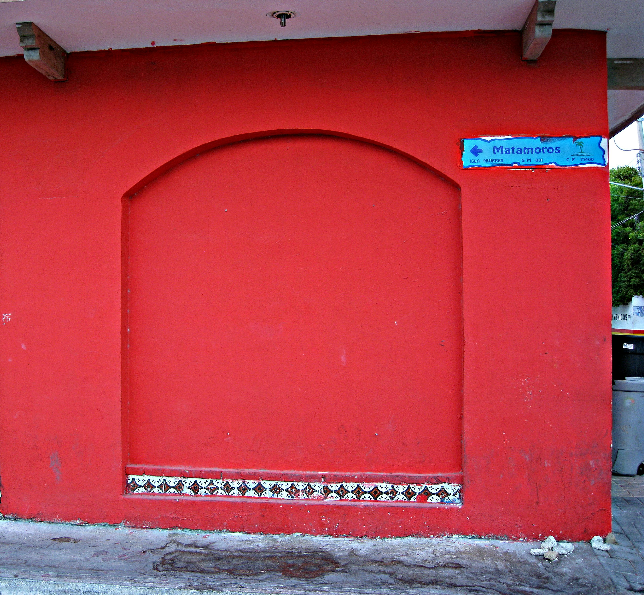 Bright red wall featuring an empty arch and decorative tile border, with a street sign indicating Matamoros. The simplicity highlights the bold color and architectural elements.