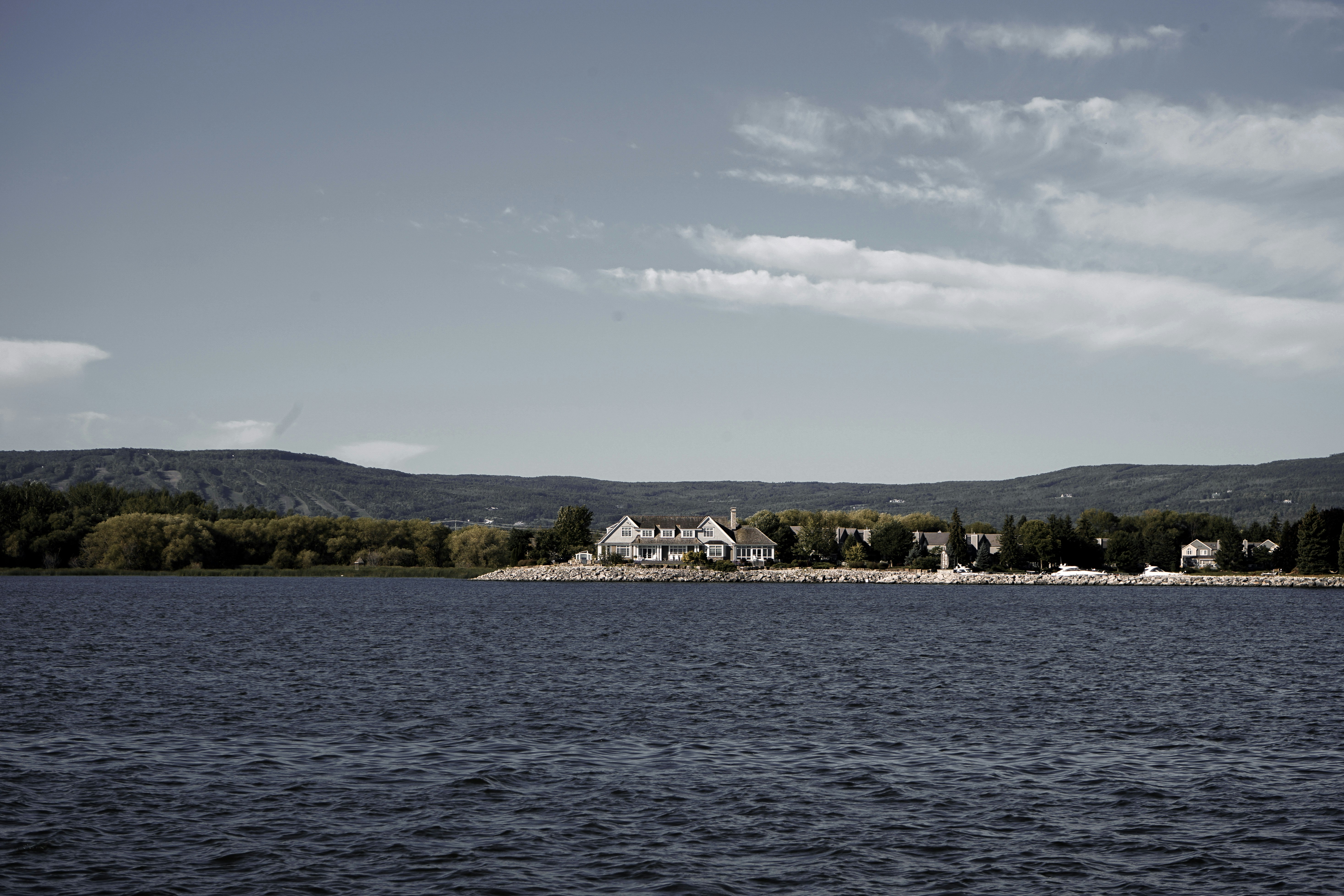Distant view of a house by a calm lake with hills and clouds in the background.