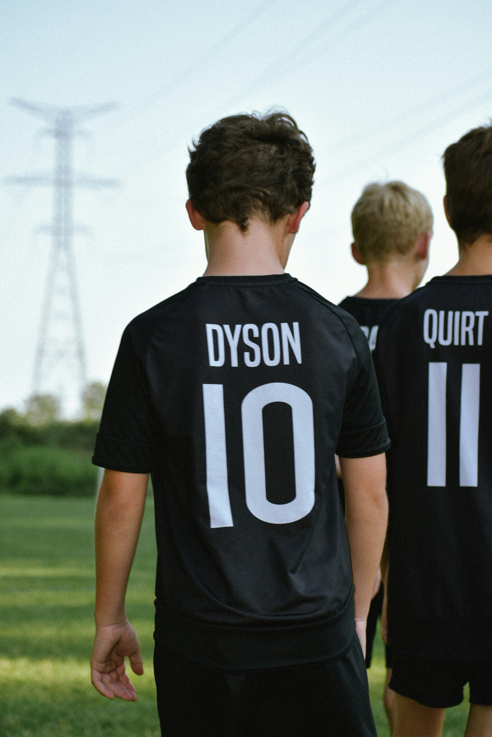 Backs of three players in black jerseys with names 'DYSON' and 'QURT' on a sunny field