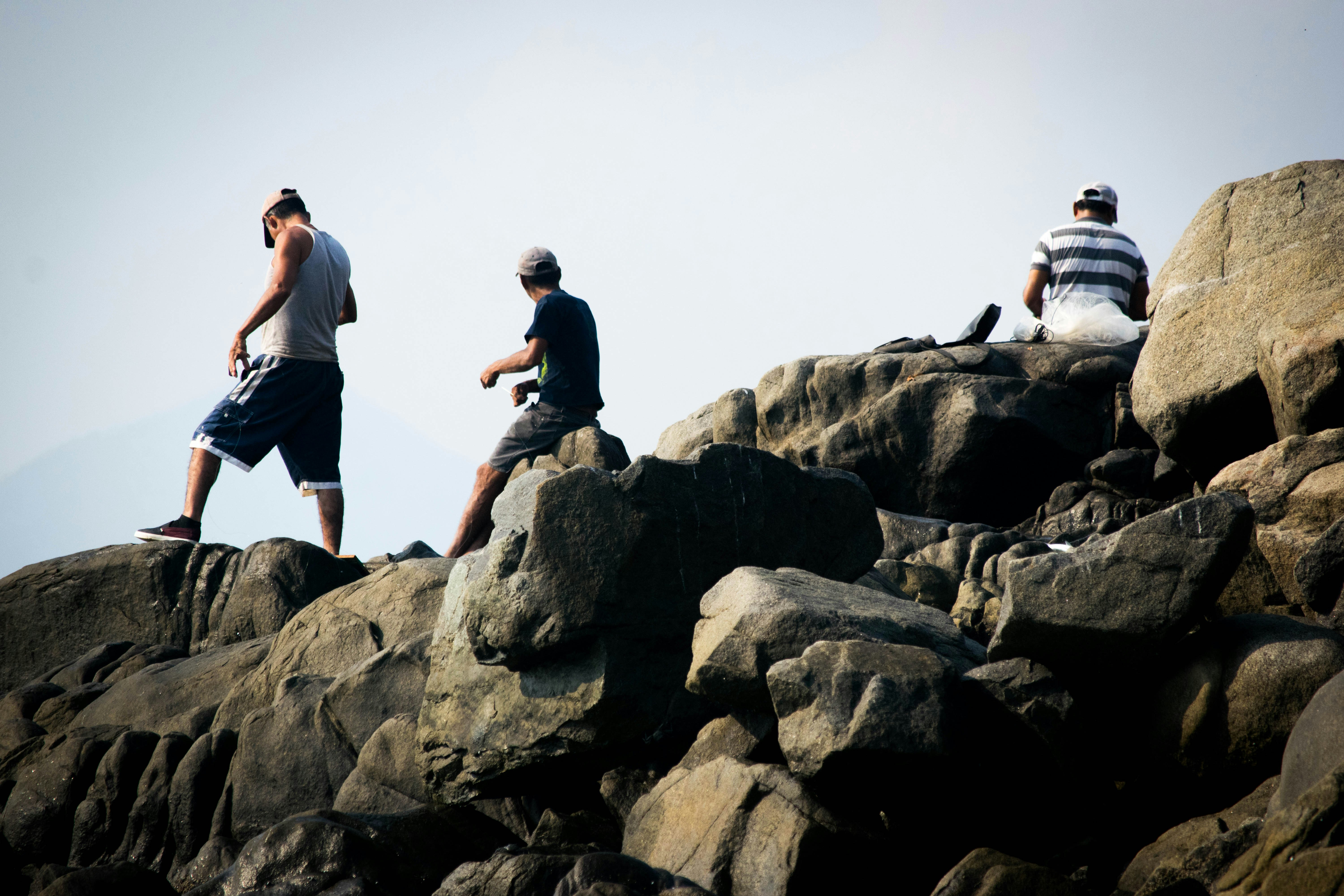 Three fishermen navigate large, rugged rocks by the sea under a clear sky.