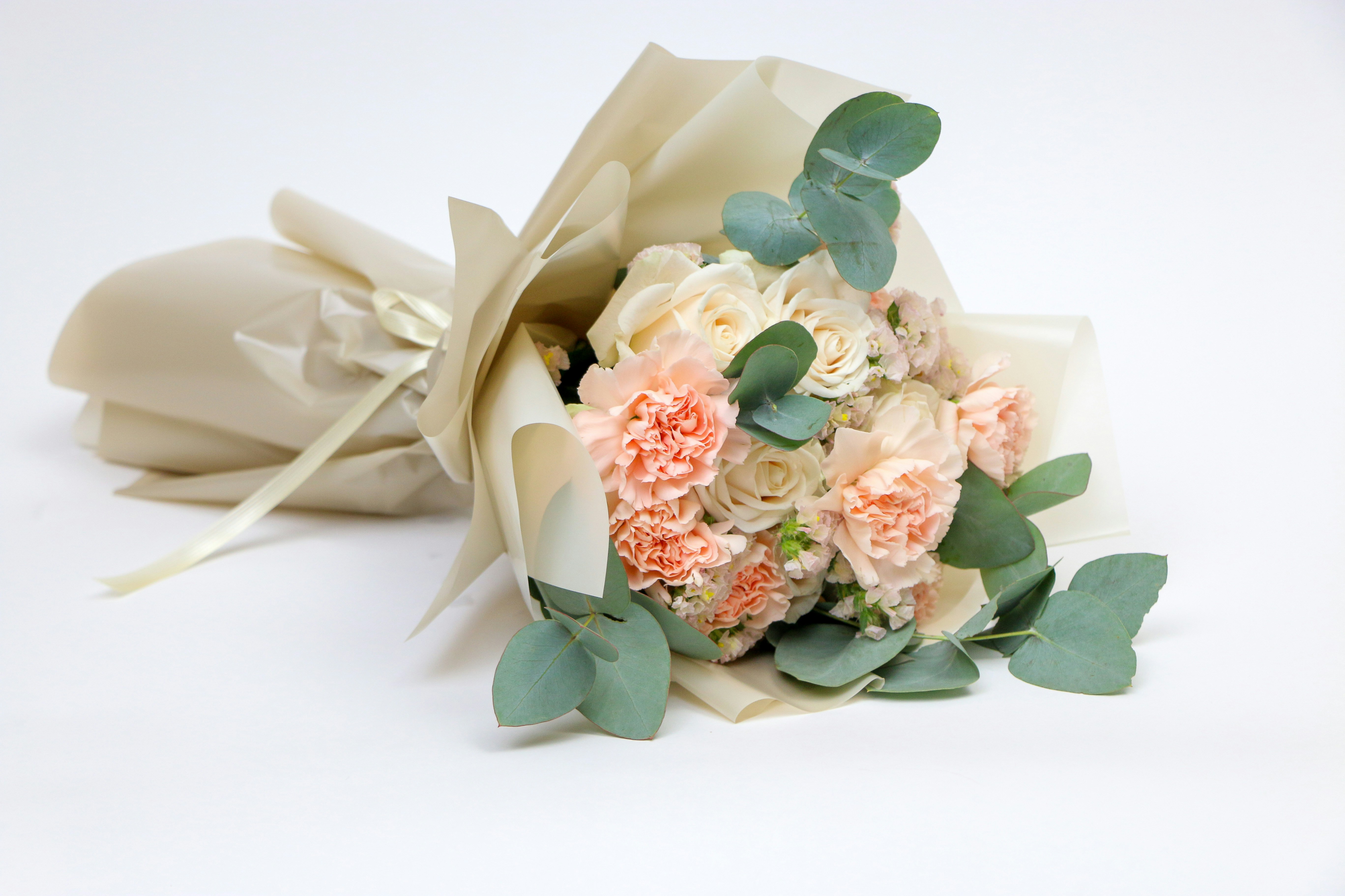 Bouquet featuring soft peach carnations and white roses, elegantly wrapped in cream-colored paper with eucalyptus leaves peeking through.