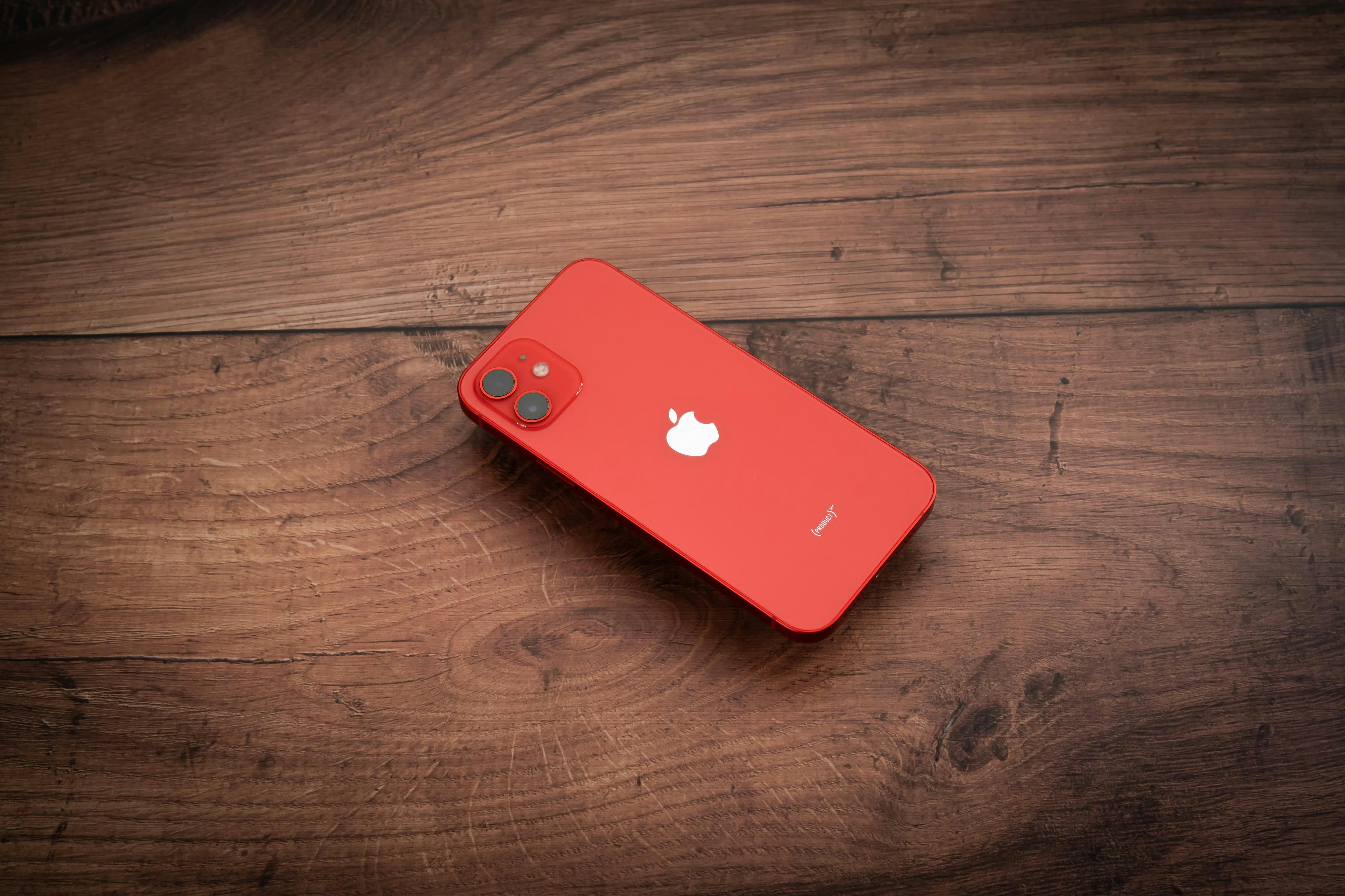 A sleek red Apple smartphone resting on a wooden surface, showcasing its modern design and logo. The warm tones of the wood complement the device's vibrant color.