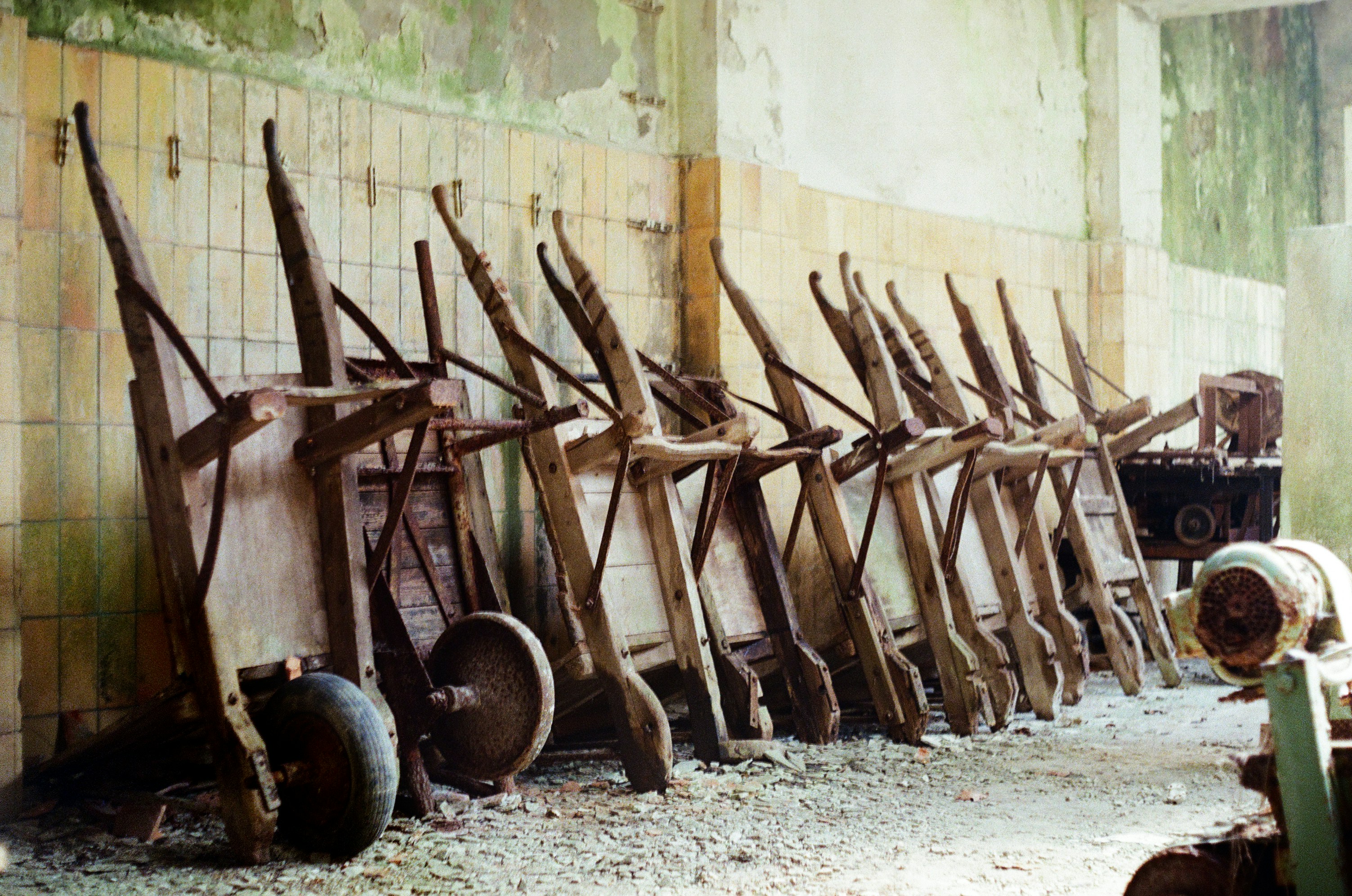 Row of antique wheelbarrow frames lean against a peeling green wall inside a dusty, abandoned workshop.