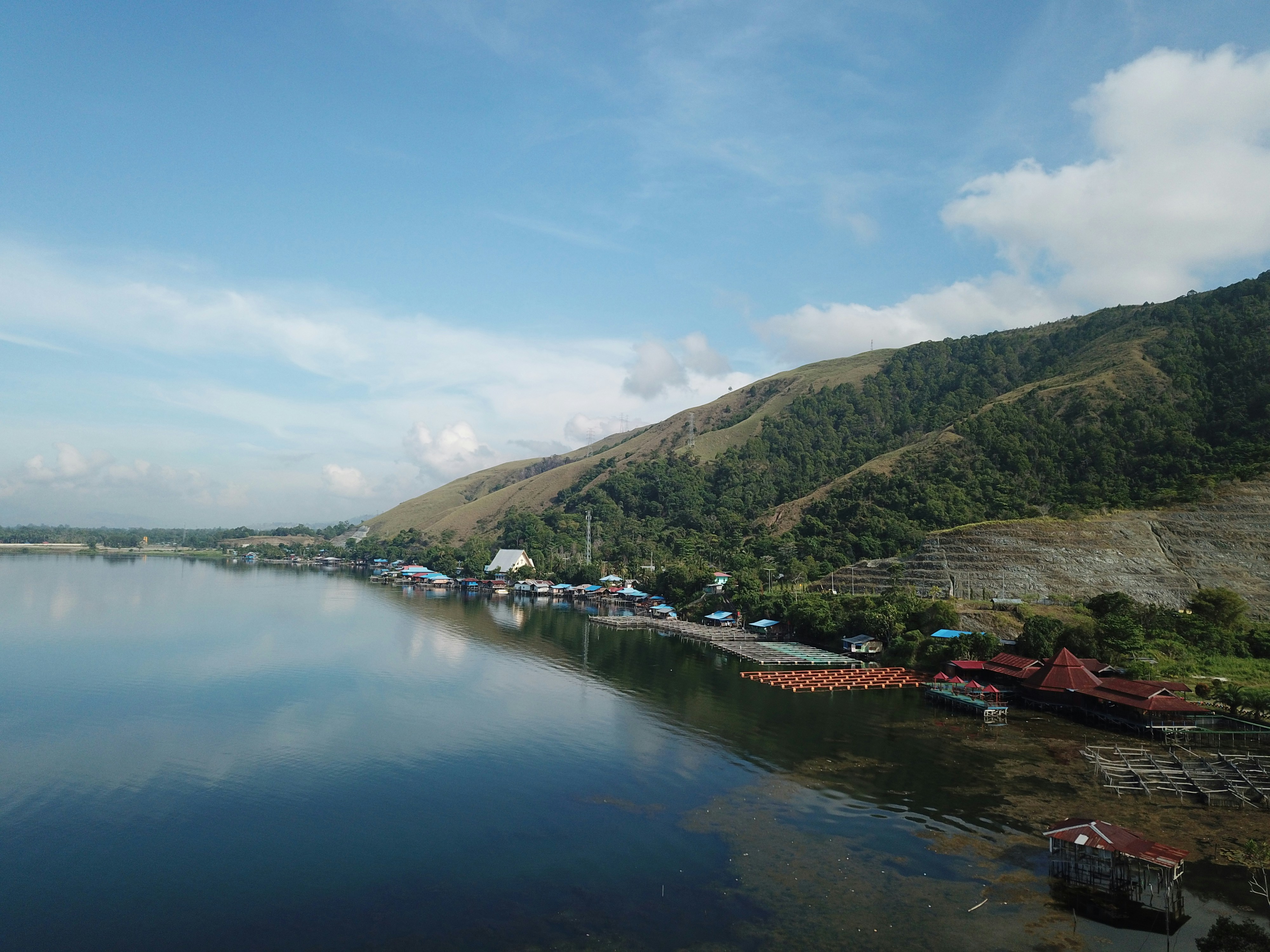 Tranquil lake reflecting rolling hills and scattered buildings under a clear blue sky.