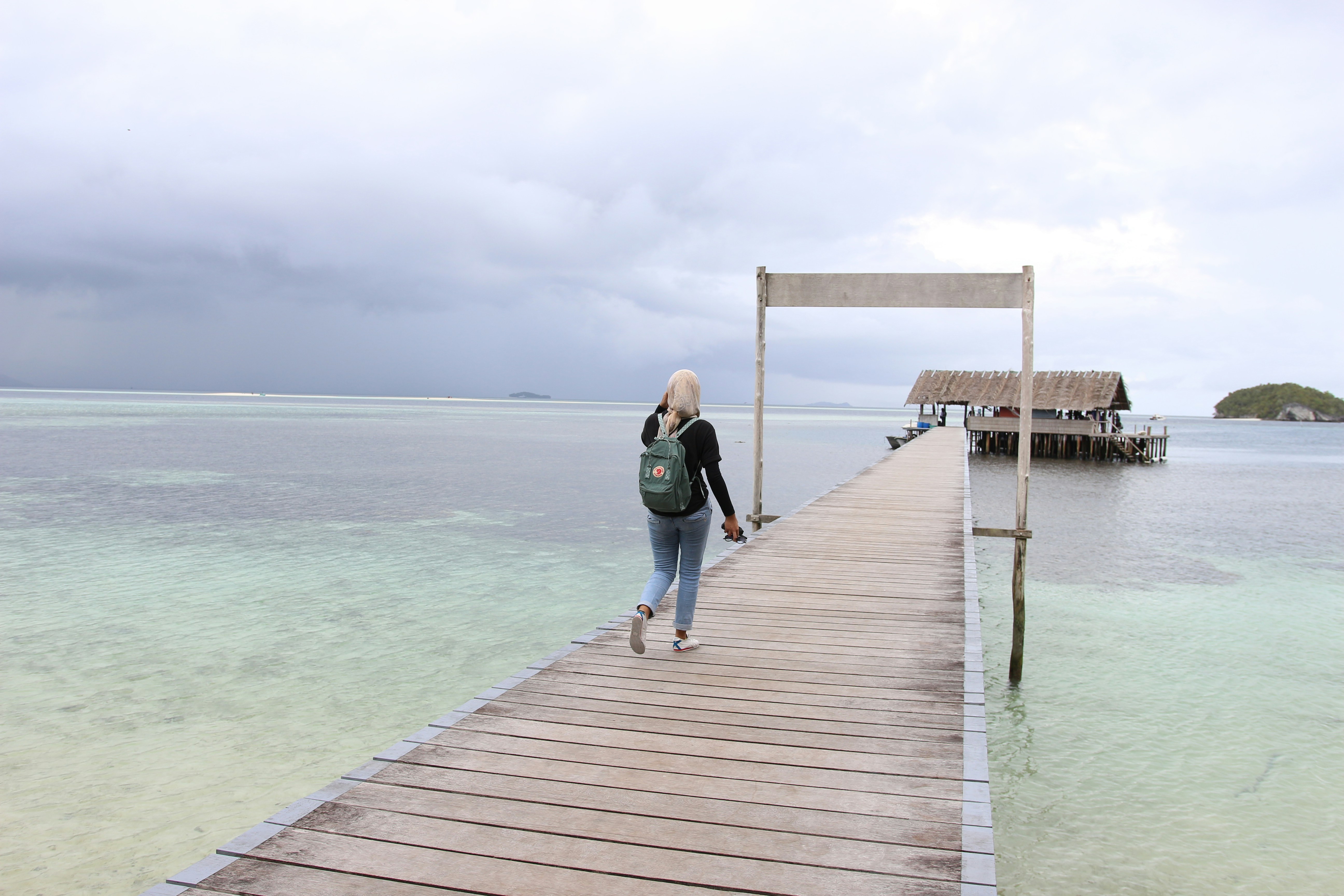 Person walking on a wooden pier extending over clear turquoise waters toward a thatched-roof hut.