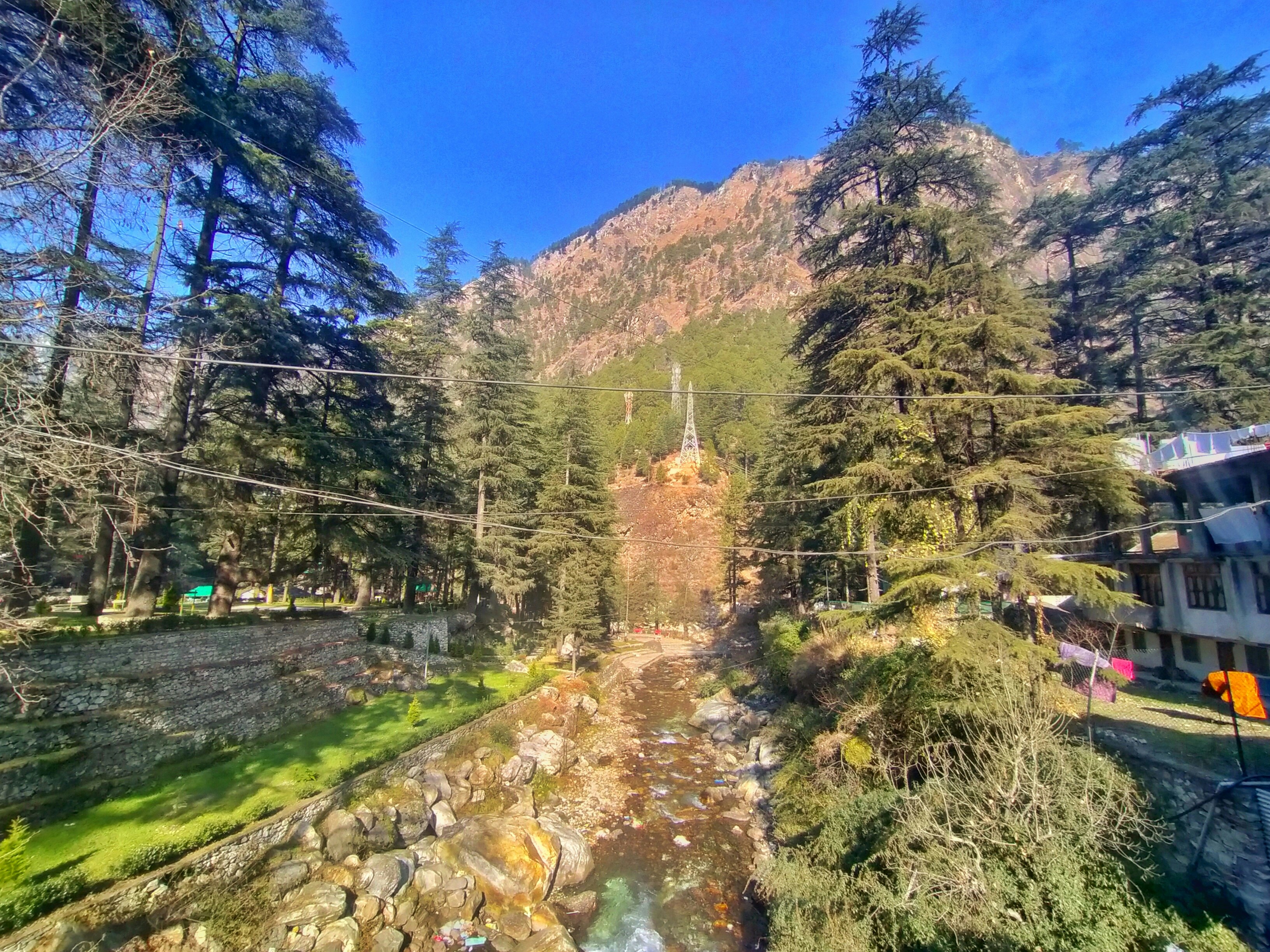 Photograph of a sunlit river valley framed by pines, with a church spire rising on a hill in the distance.