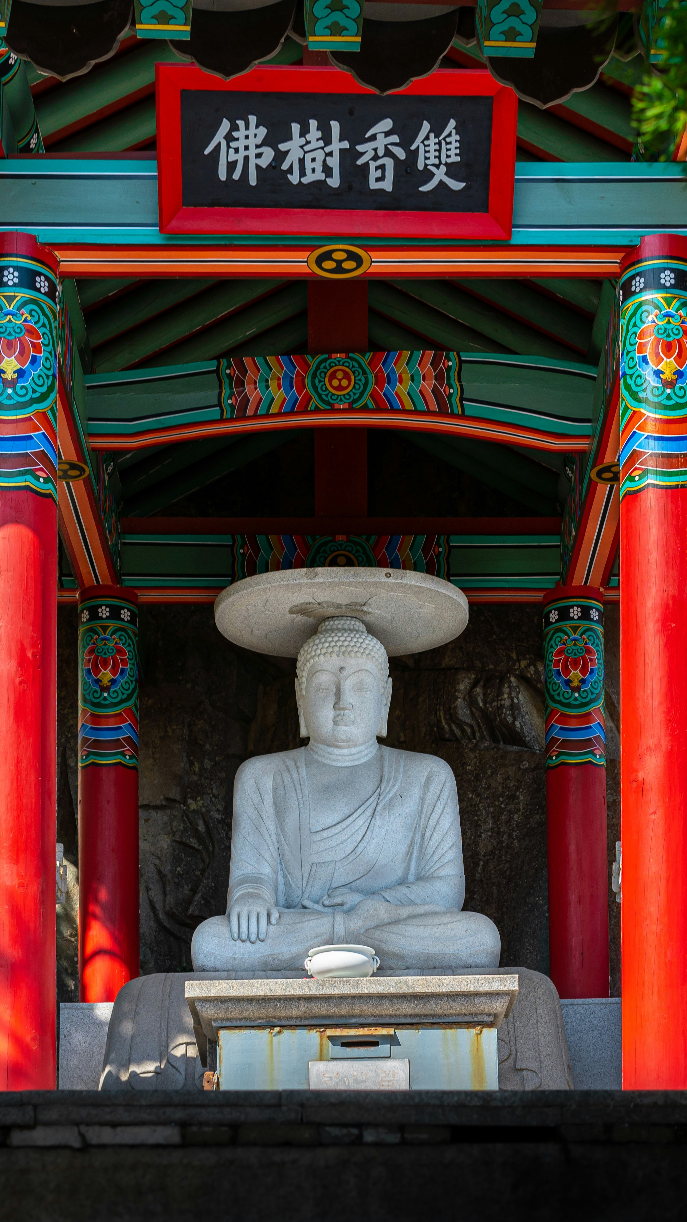 White stone Buddha statue seated beneath a vibrant, intricately designed pavilion, surrounded by colorful pillars. A tranquil atmosphere invites reflection.