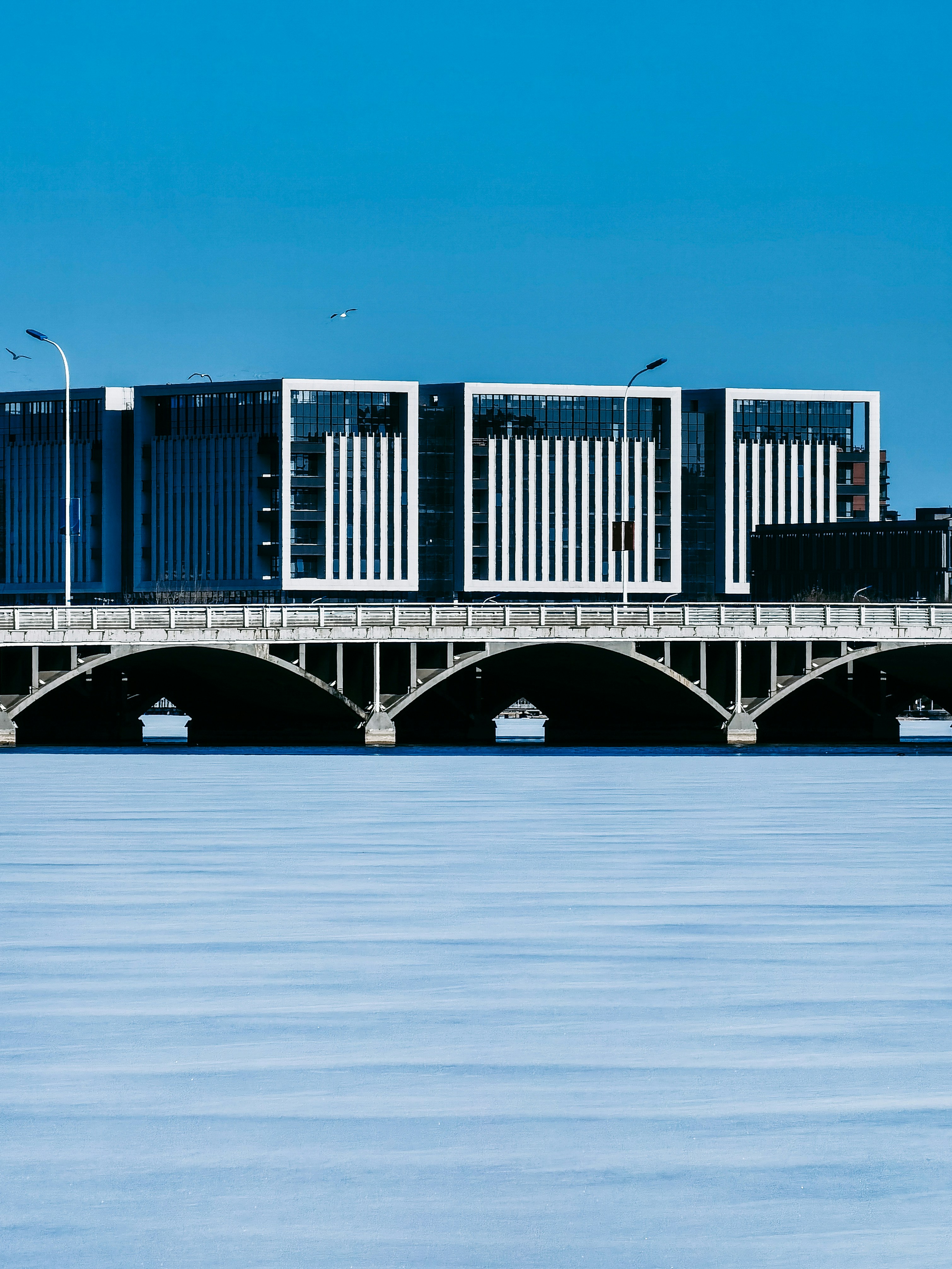 Daytime photograph of a bridge with arched supports spanning a calm blue river, backed by a row of modern glass-fronted buildings under a clear sky.