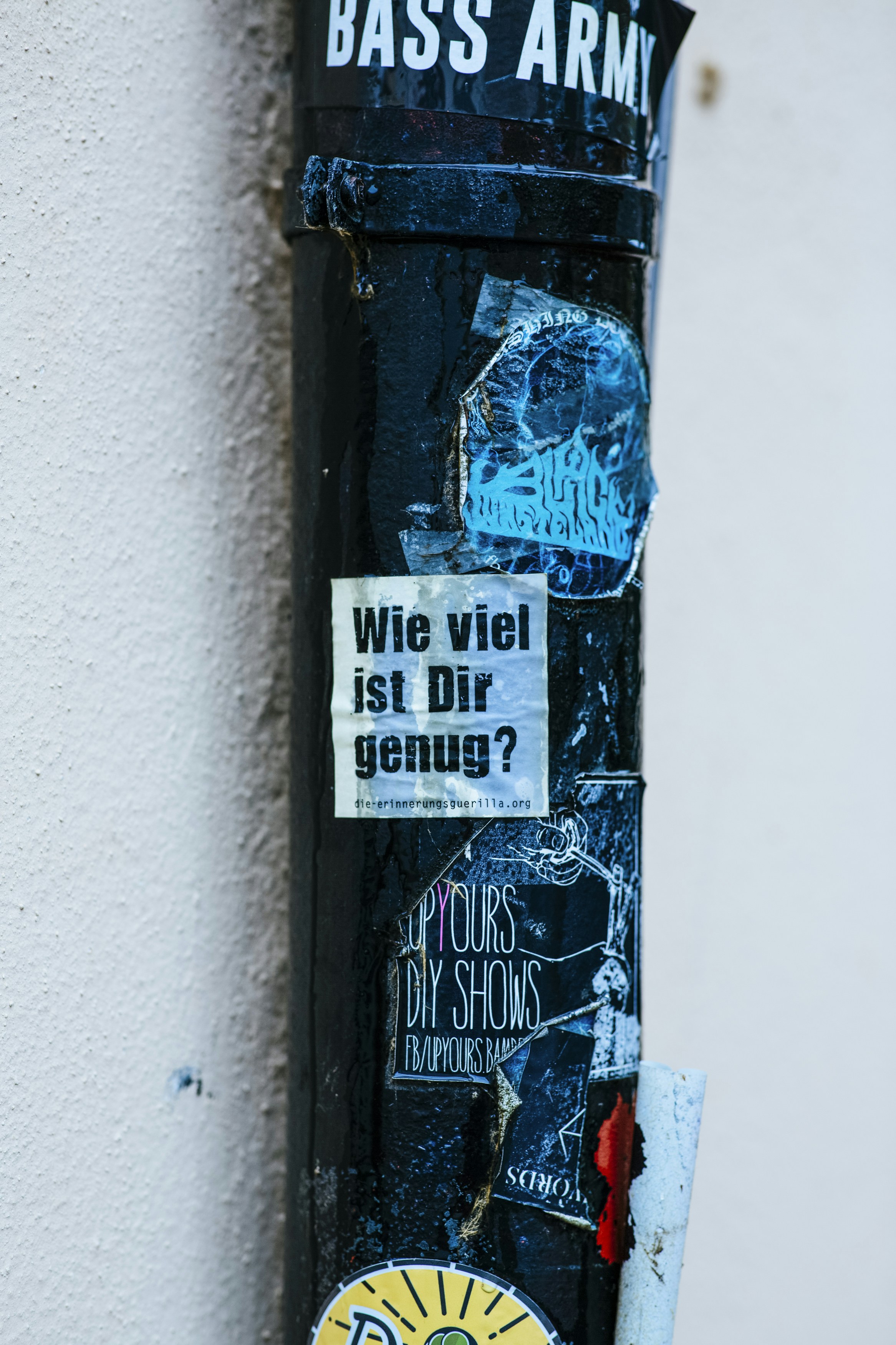 A weathered pole adorned with various stickers, prominently featuring the question 'Wie viel ist Dir genug?' in bold lettering.