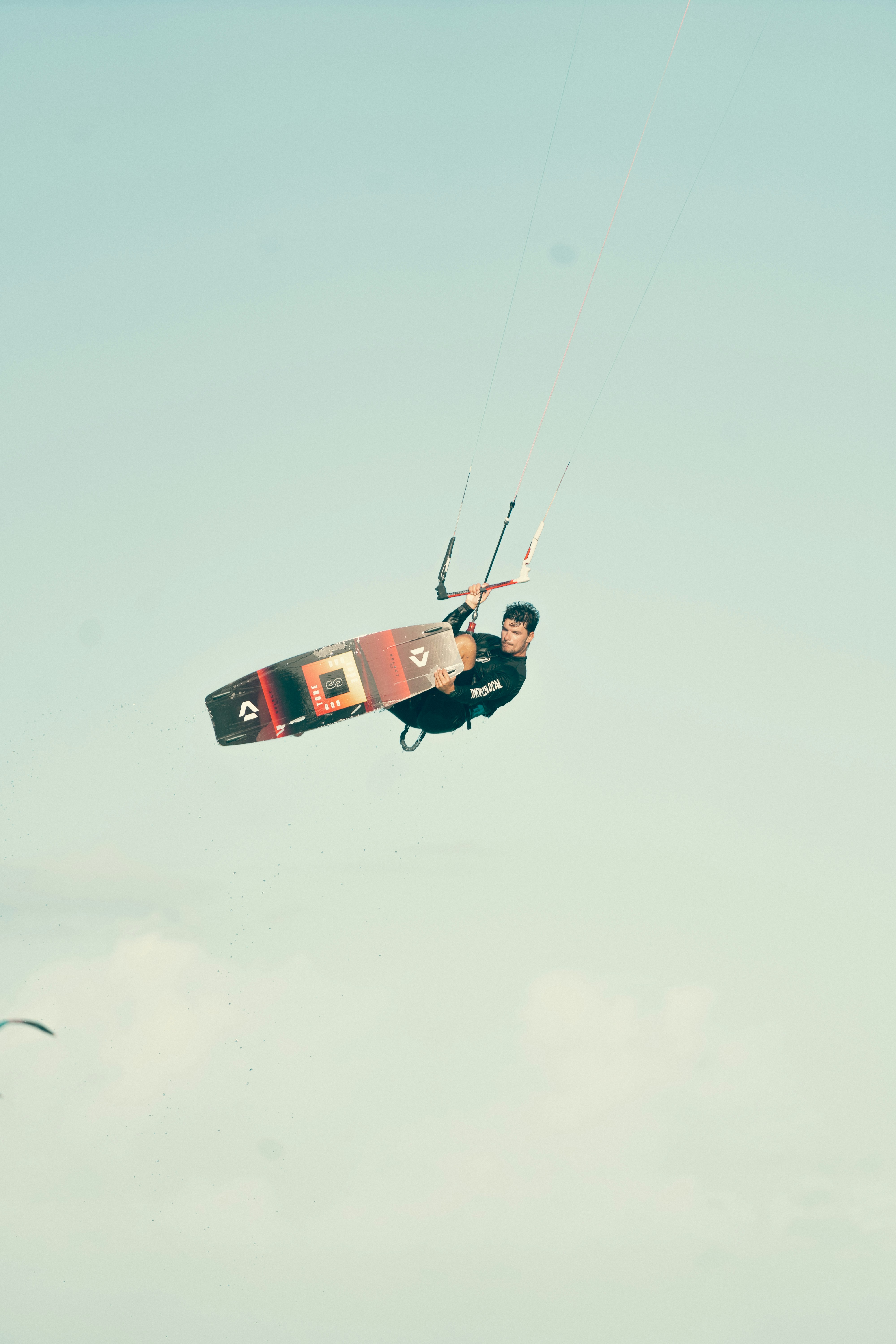 A kiteboarder performs an aerial maneuver against a clear sky, showcasing skill and athleticism. The image captures the dynamic motion and excitement of the sport.