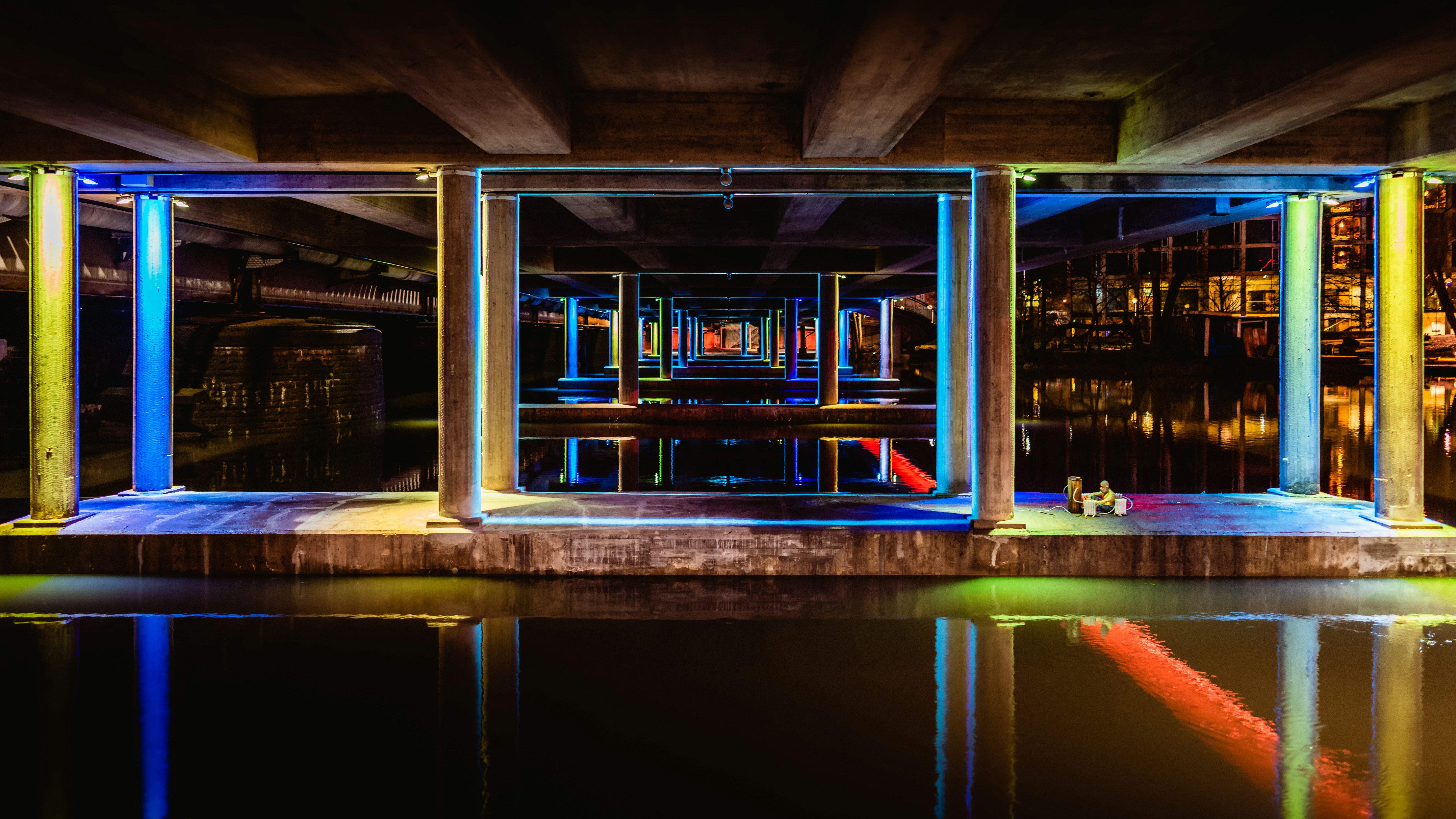 Colorful illuminated beams and their reflections under a bridge at night.