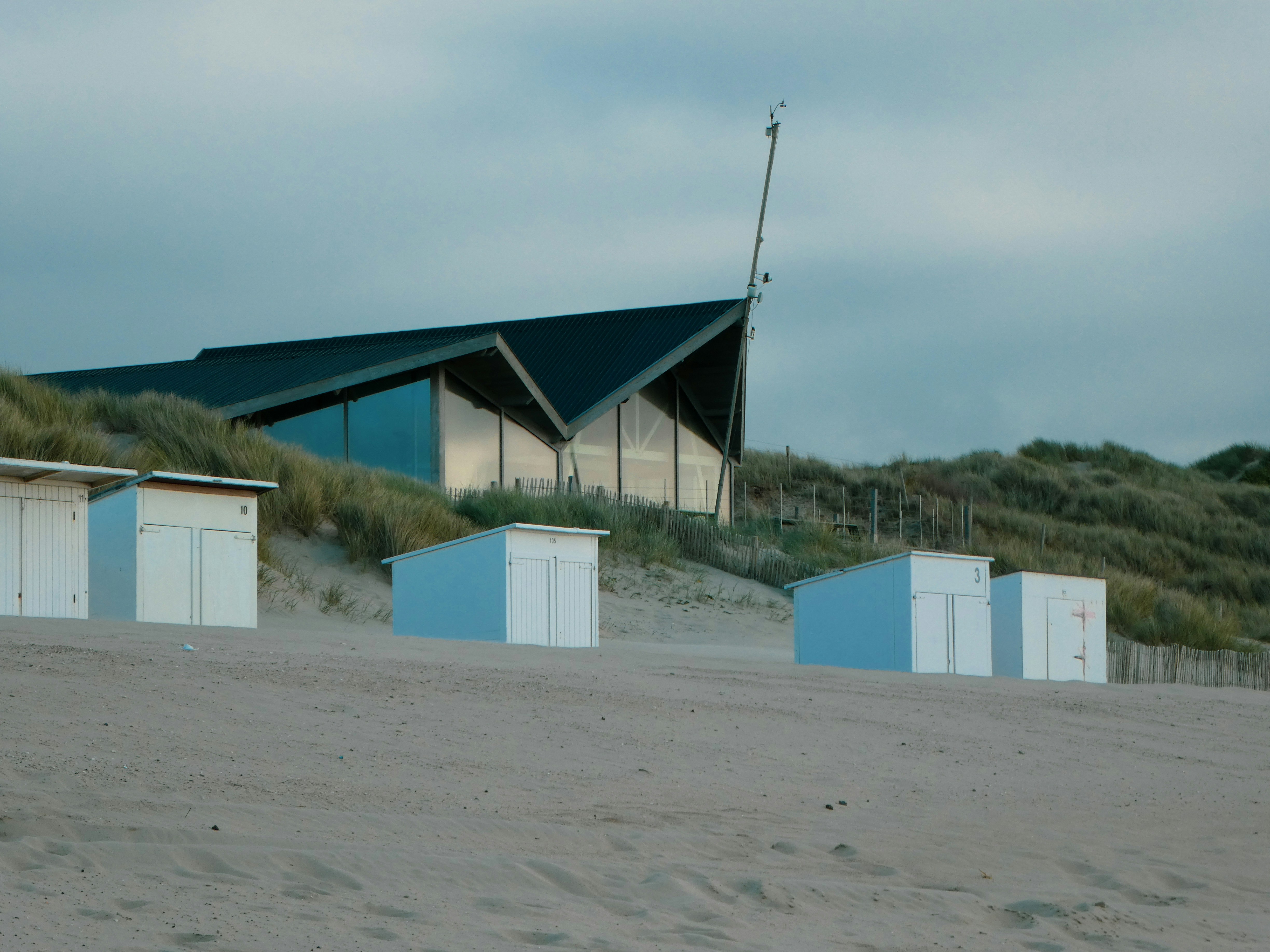 Contemporary beach house set against a backdrop of sand dunes, with minimalist beach huts in the foreground. 