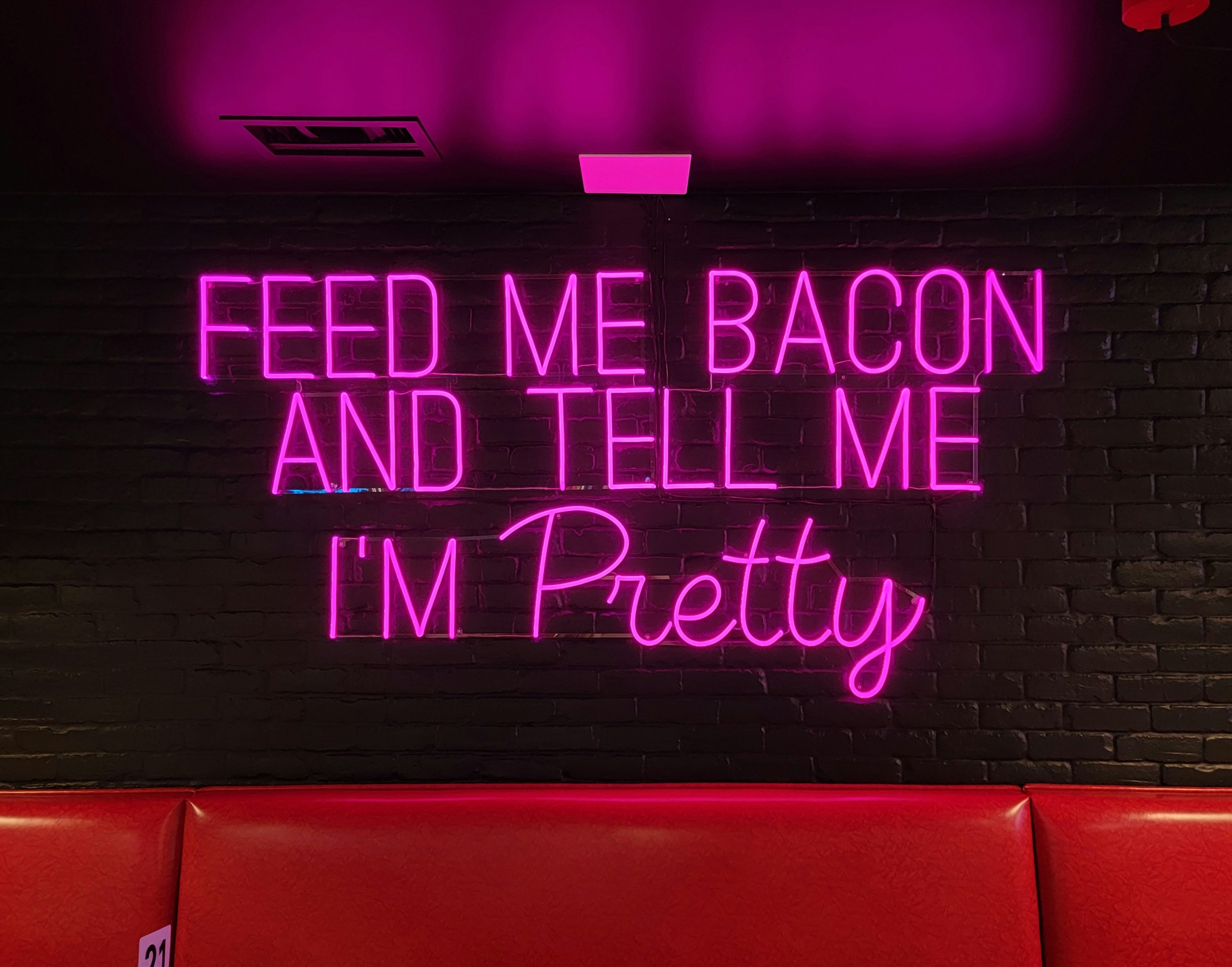 Pink neon sign reads 'FEED ME BACON AND TELL ME I'M Pretty' on a dark brick wall above a red booth.