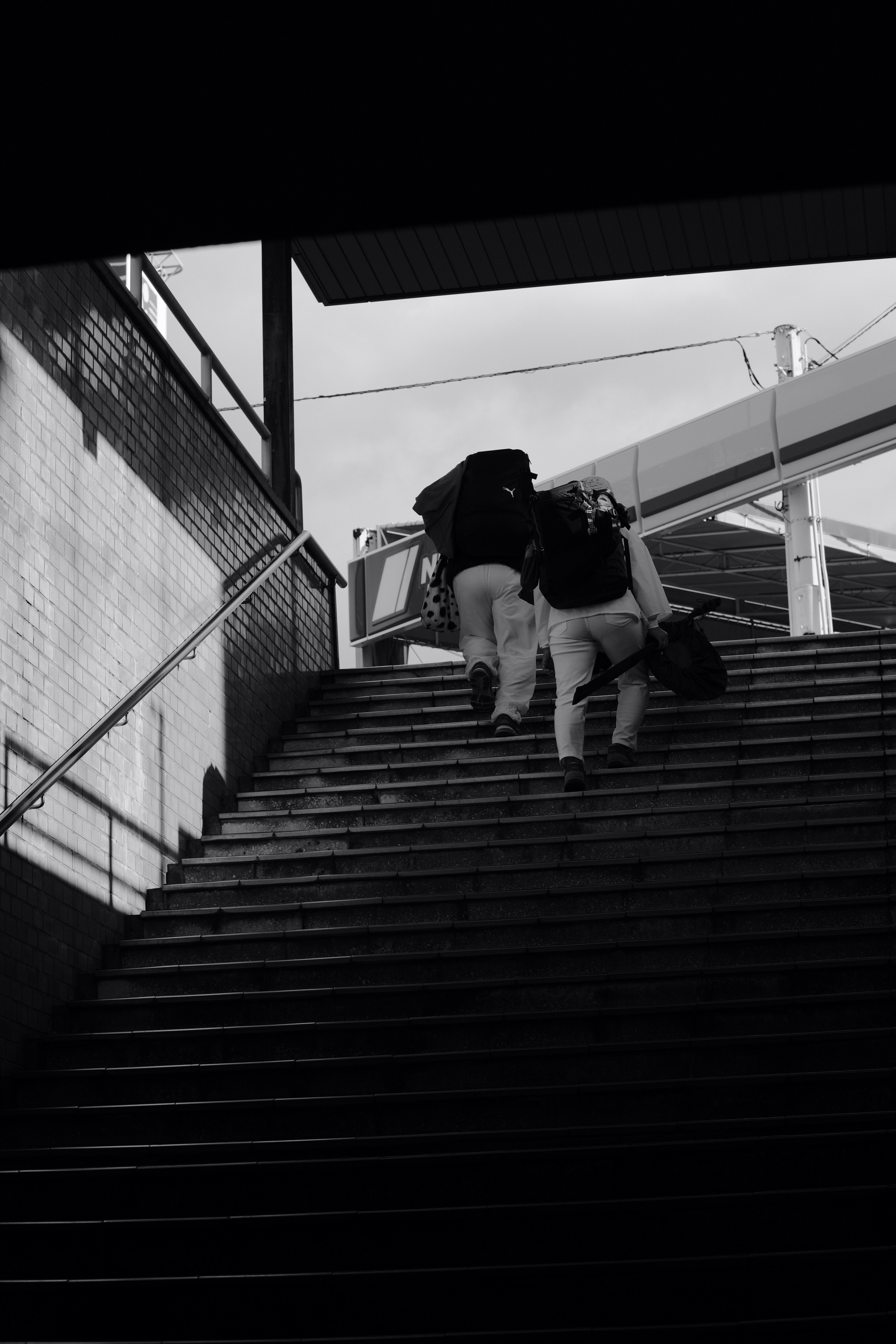 Monochrome Ascent: Two Travelers with Large Backpacks Climb Transit Stairs