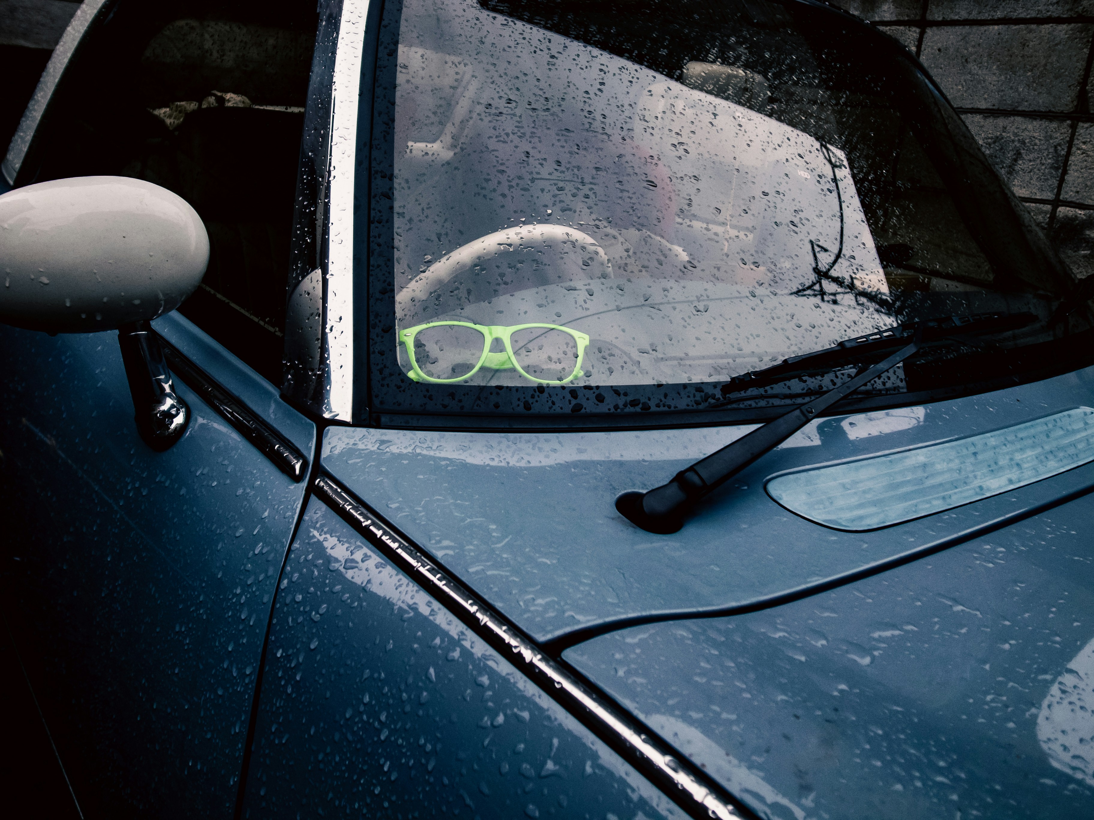 Neon-green sunglasses rest on the rain-speckled dashboard behind the windshield of a blue compact car.