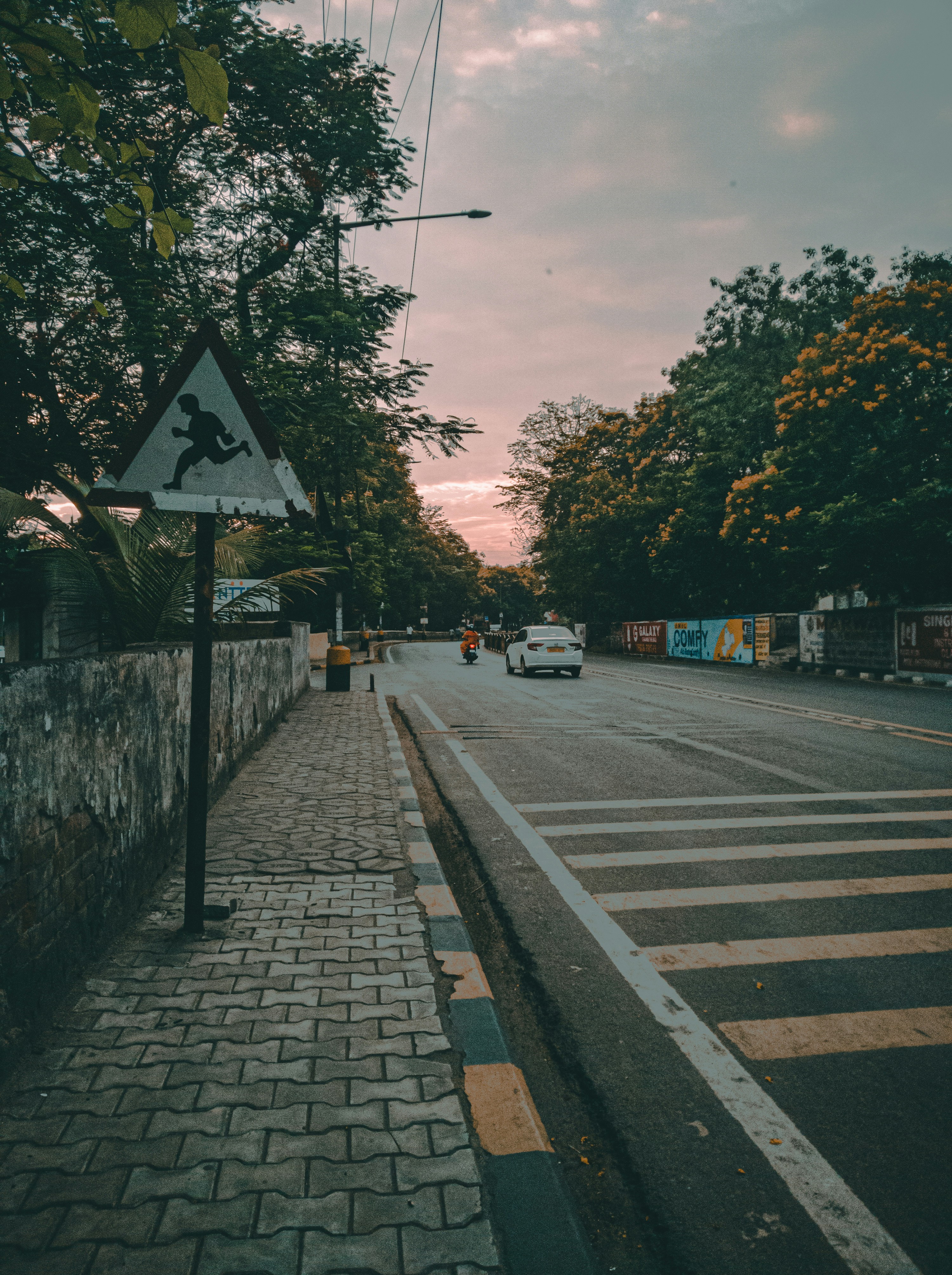 A street view featuring a pedestrian warning sign, with a car and motorcycle on a quiet road lined with trees at dusk.