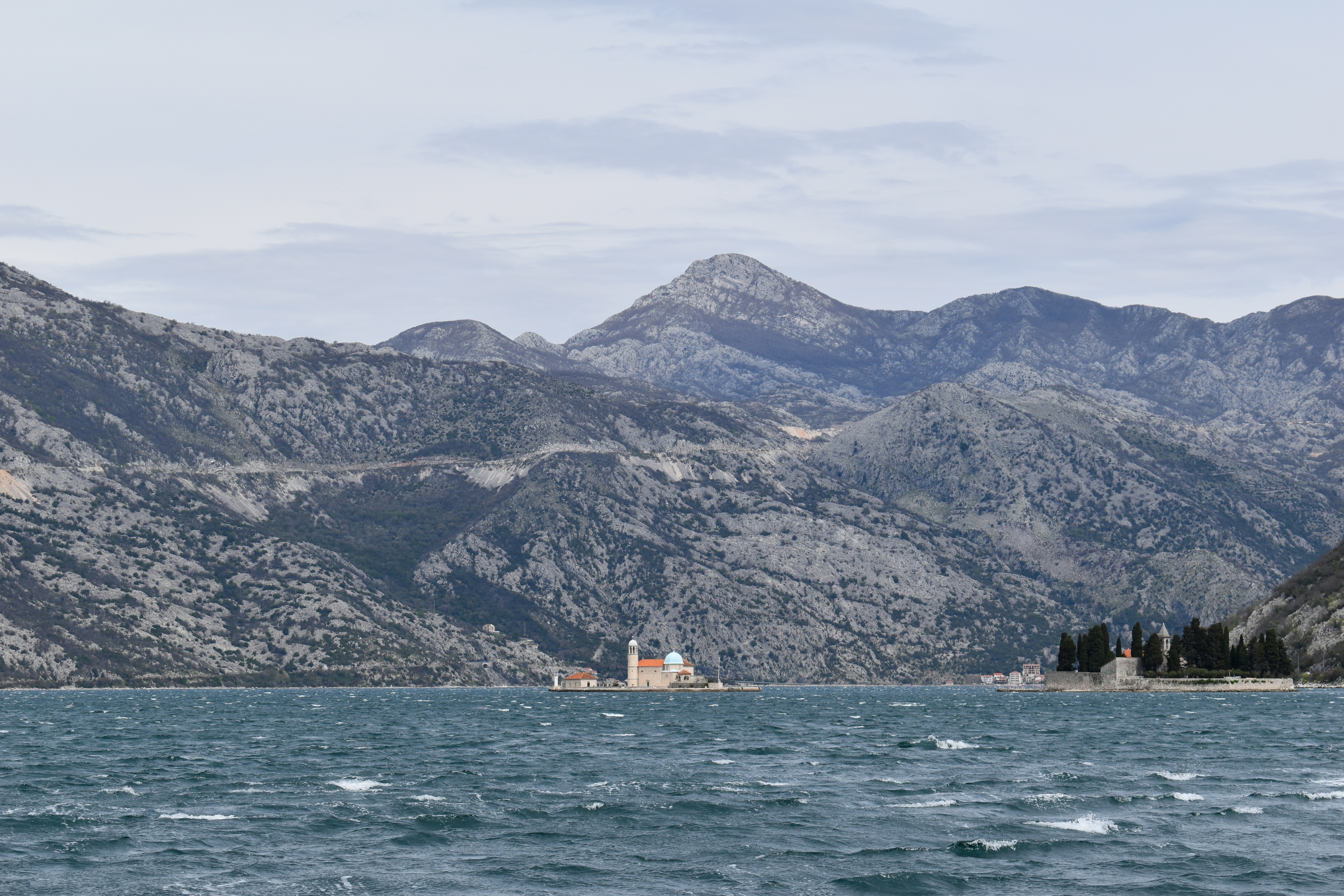 A picturesque coastal scene featuring a small island with a church set against a backdrop of rugged mountains and a choppy sea.