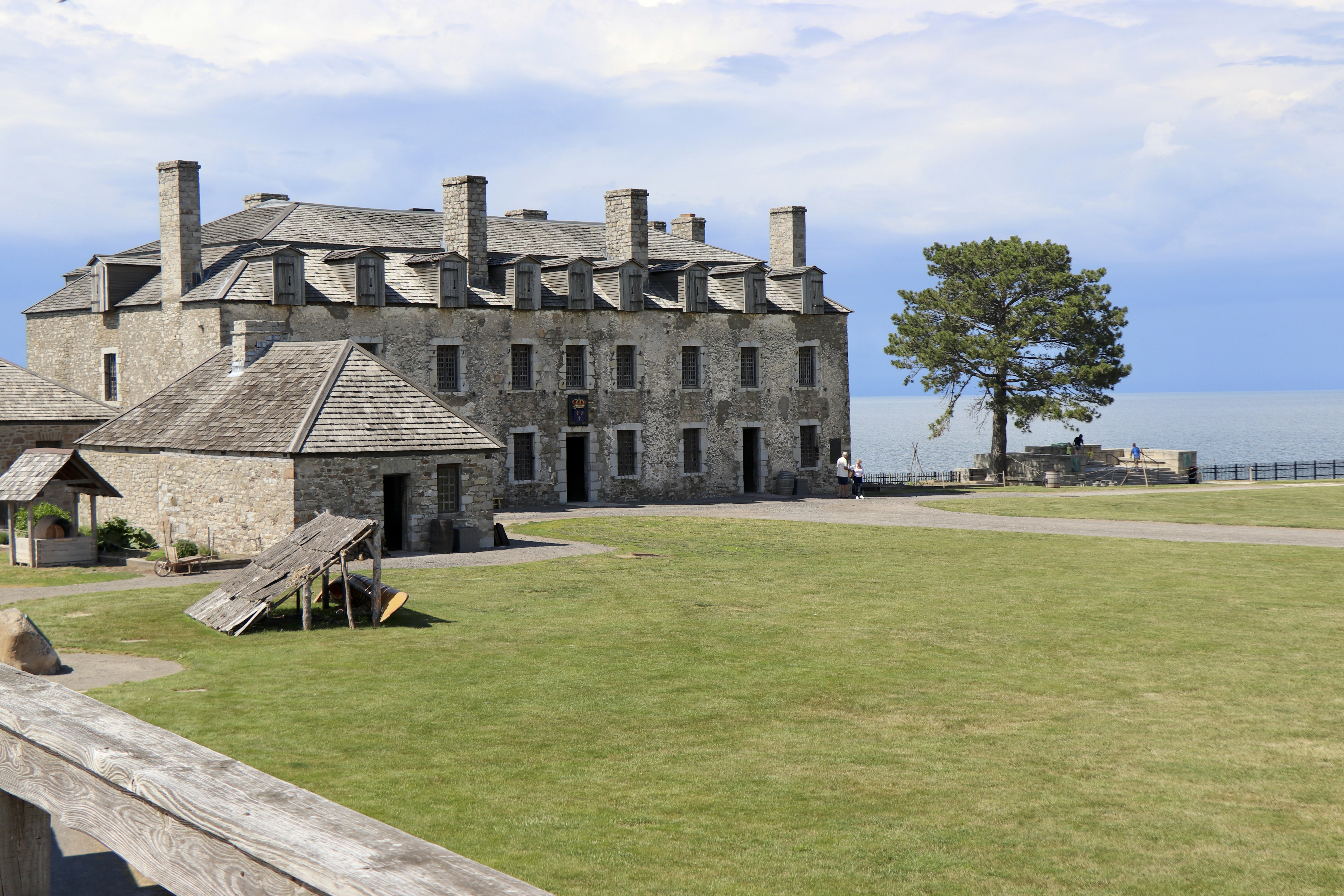 Old stone fort with multiple chimneys set against a backdrop of open grass and a distant sea under a partly cloudy sky.