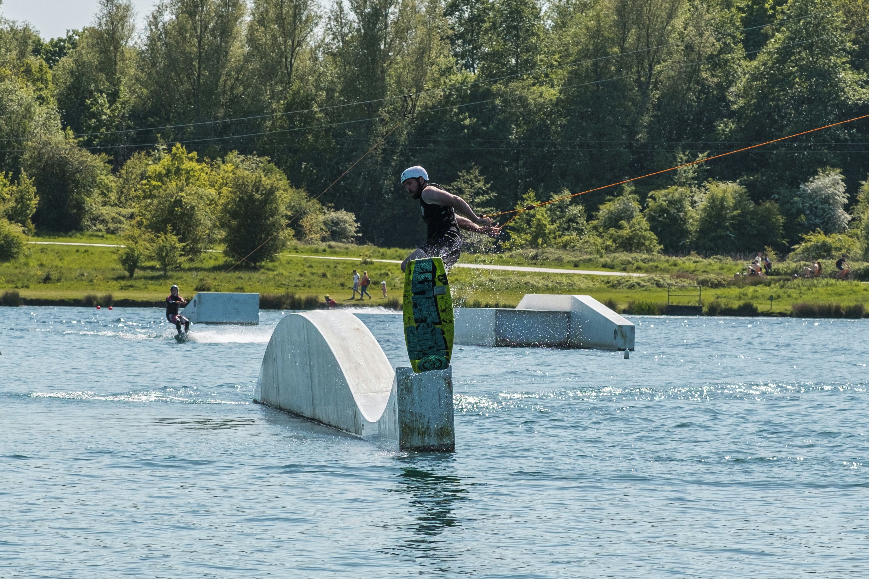 A wakeboarder executing a trick over a water obstacle, with another rider in the background. The lush greenery surrounding the lake adds vibrancy to the scene.