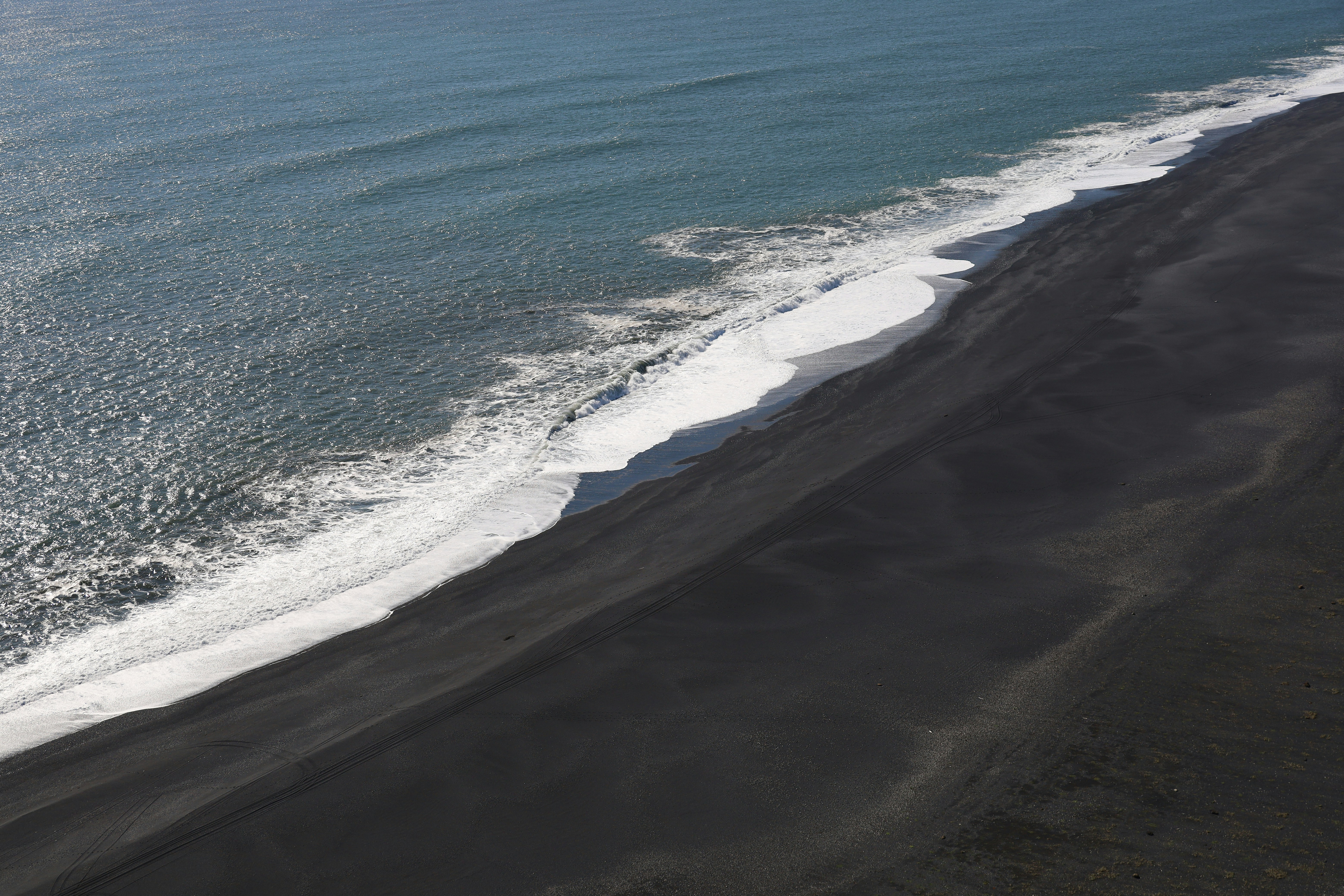 Waves gently lap against a black sand beach under a clear sky.