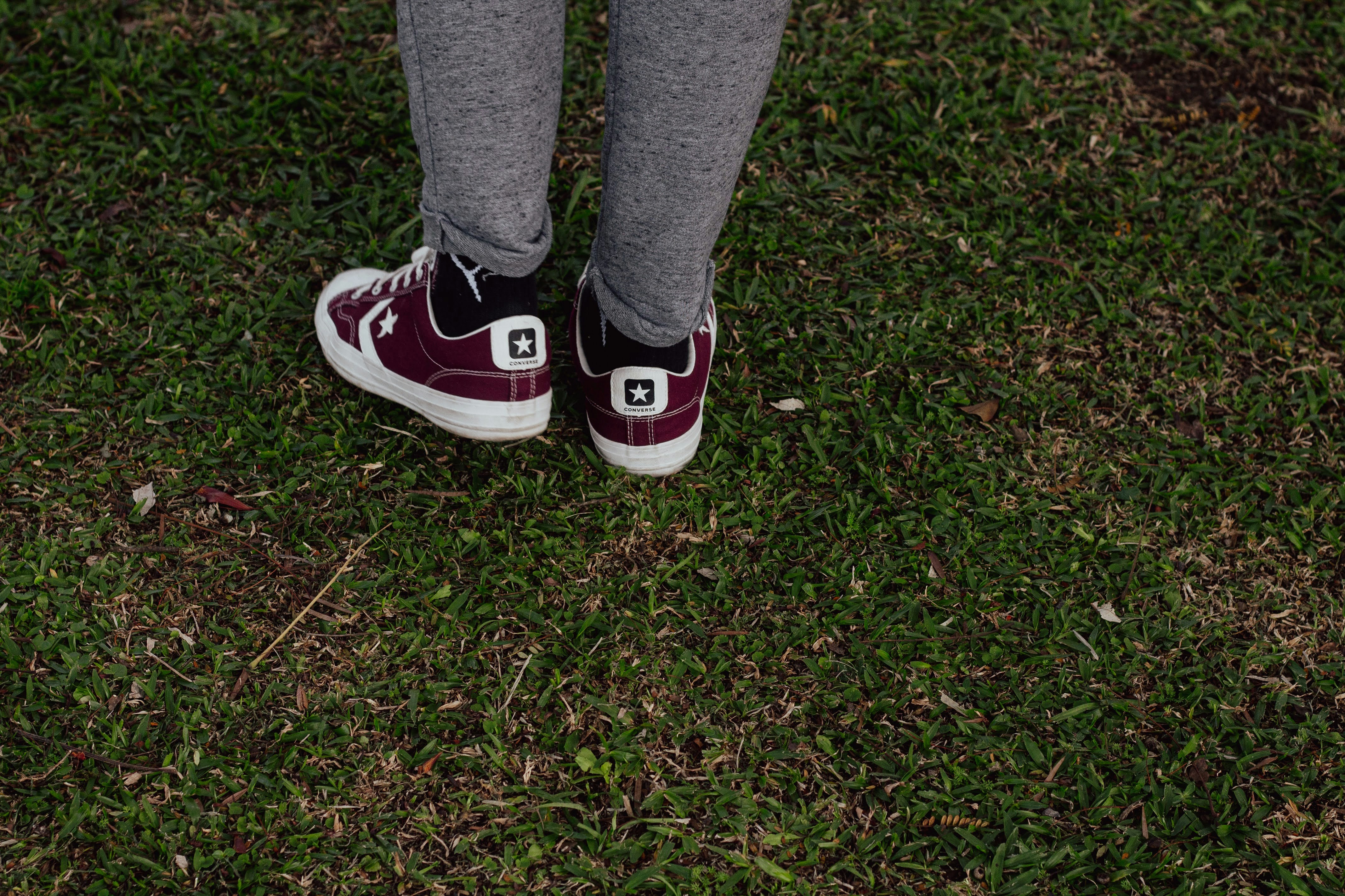 Close-up of a person’s feet wearing maroon sneakers, standing on lush green grass. The casual attire reflects a relaxed outdoor vibe.