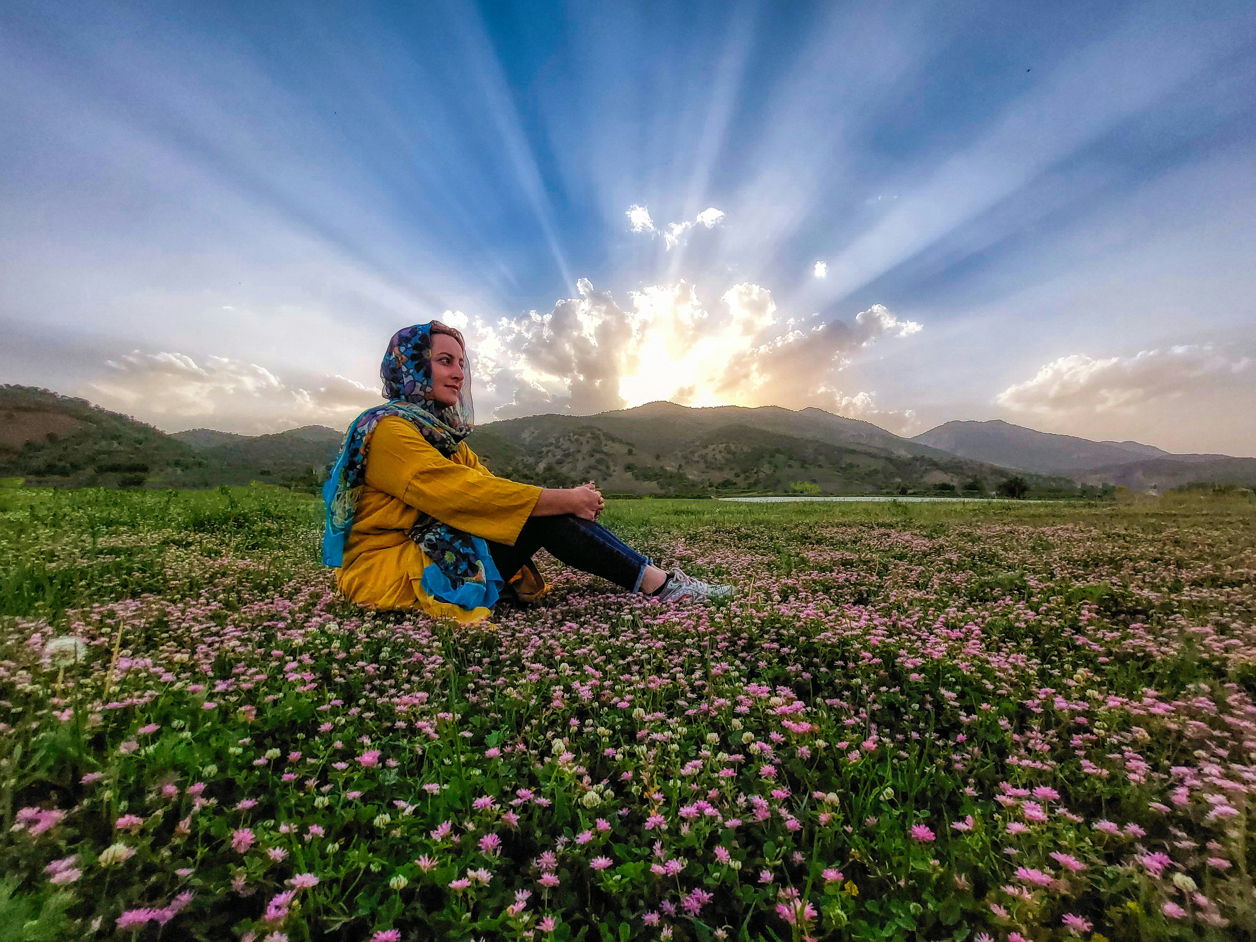Person sitting in a field of wildflowers with dramatic sun rays piercing the cloudy sky in the background.