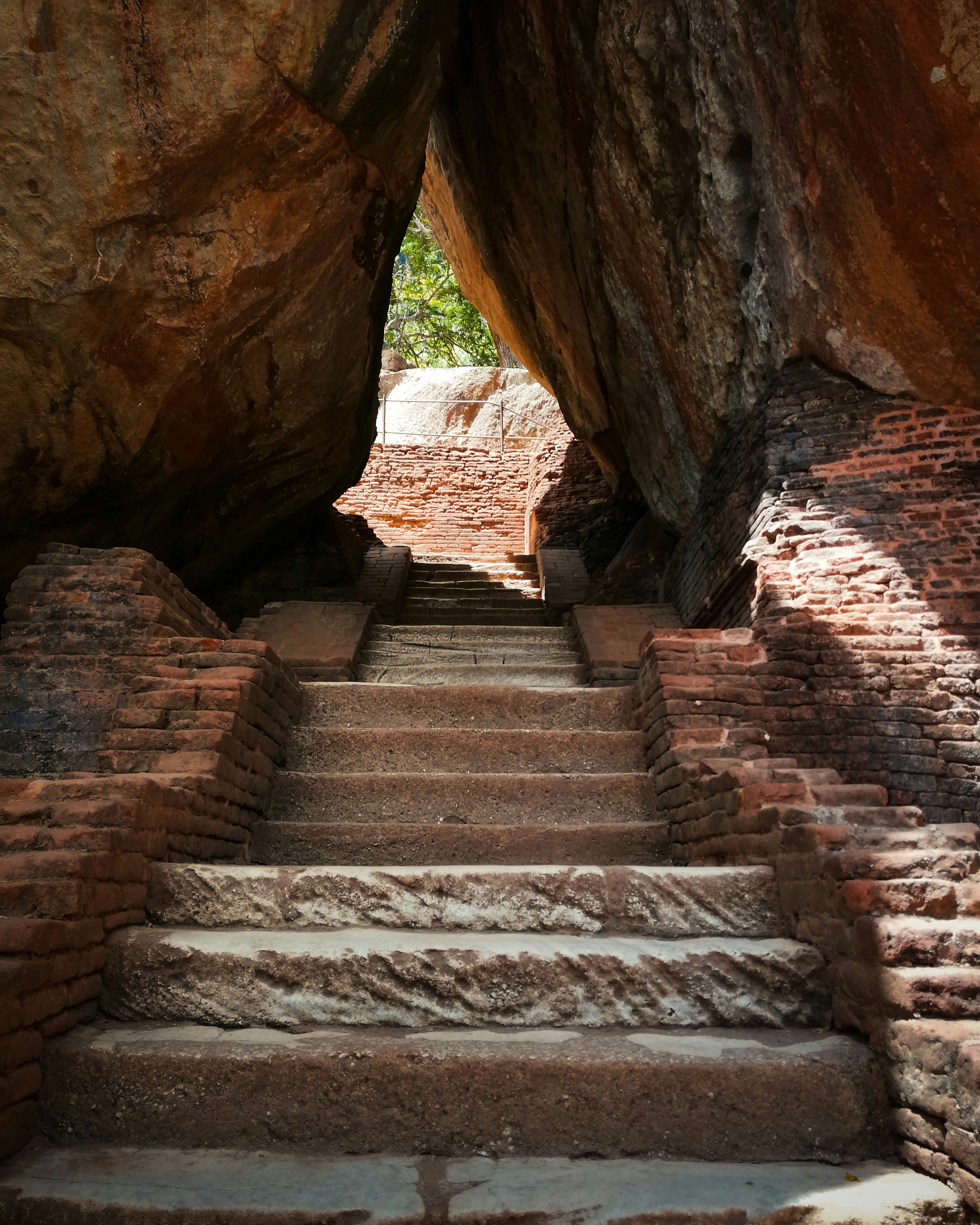Worn stone steps lead through a rocky passageway, framed by towering cliffs and dappled sunlight filtering through the foliage above.