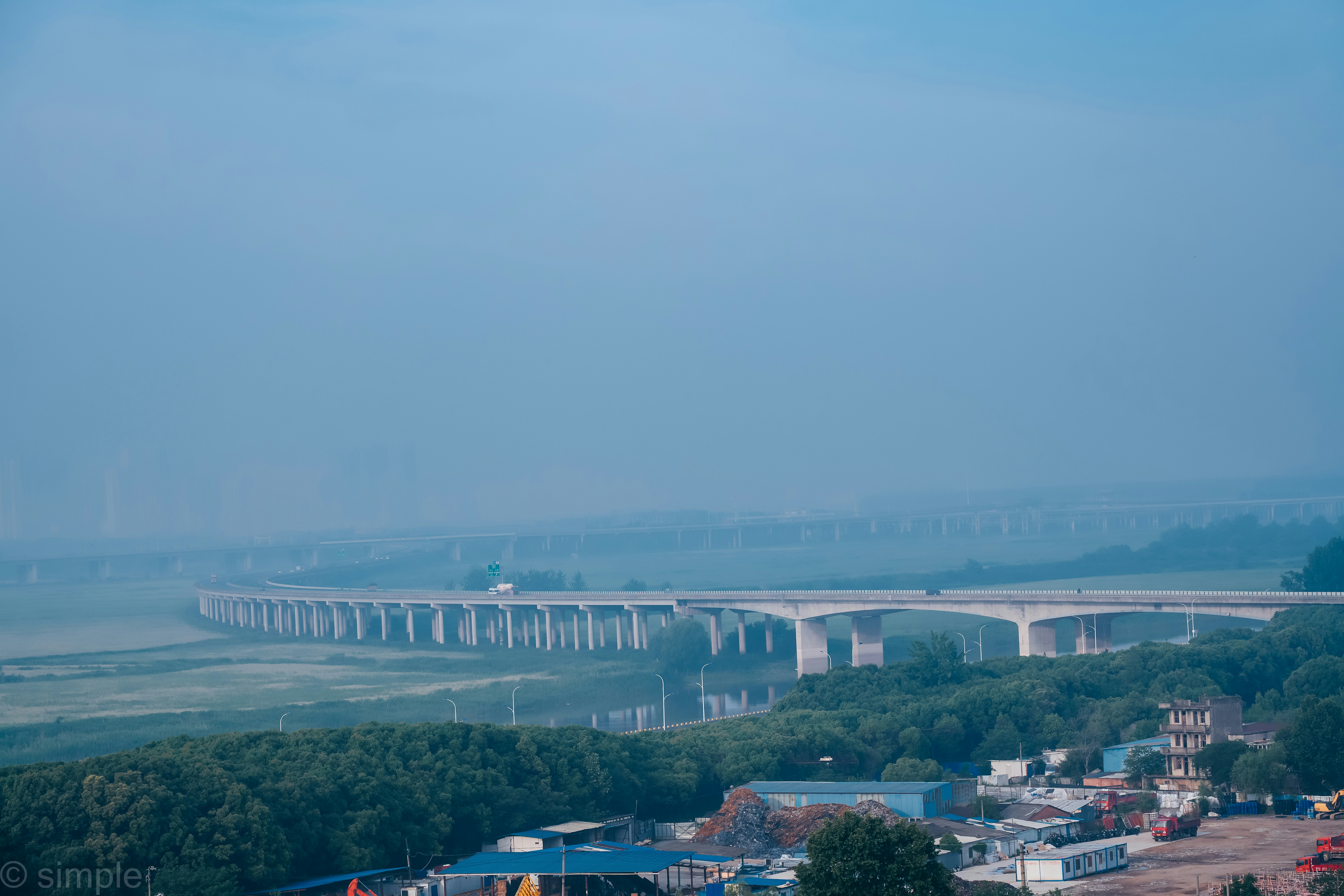 Long bridge stretching over expansive, mist-covered landscape under a blue sky.