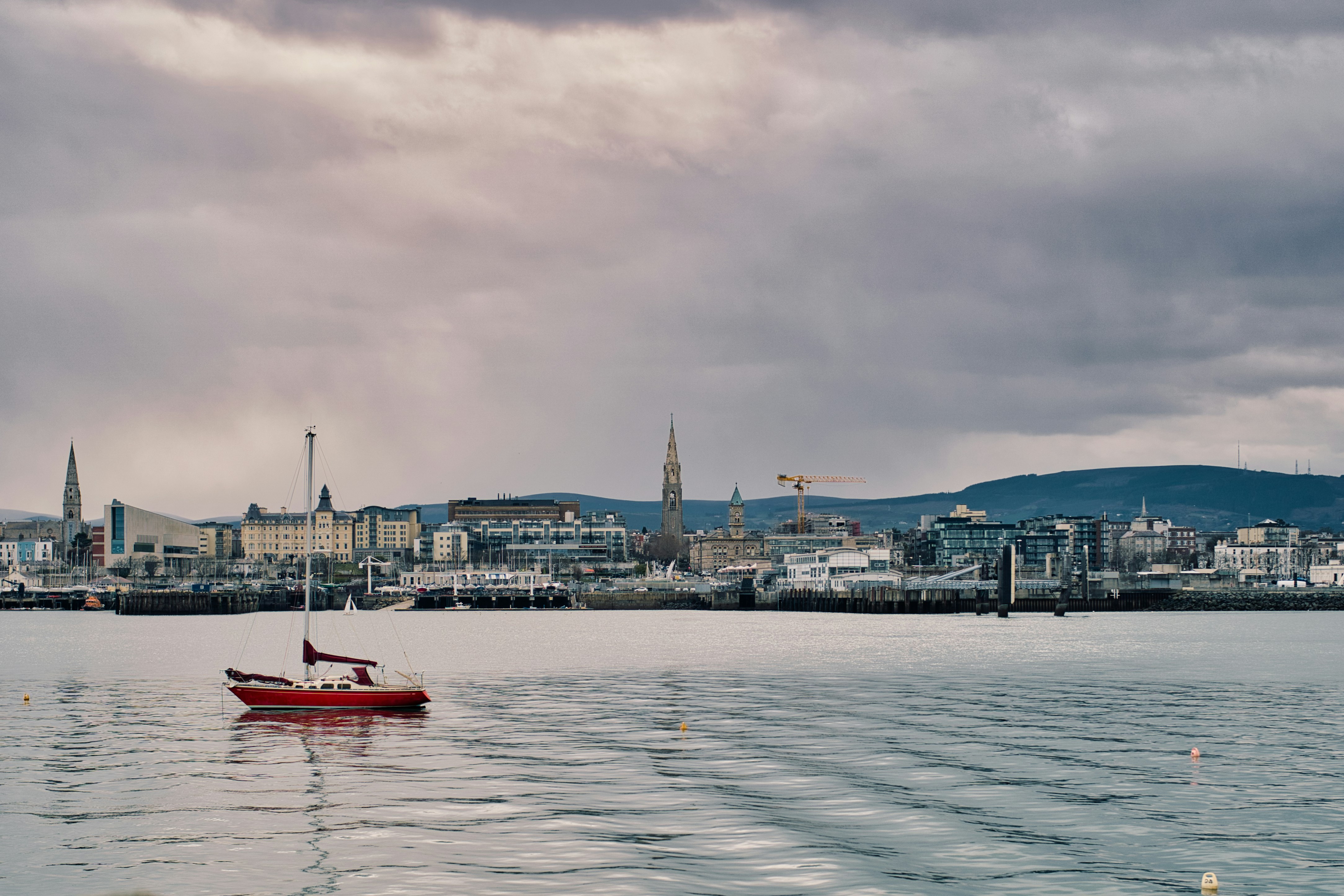 Red boat gliding across calm waters with a city skyline under a cloudy sky.