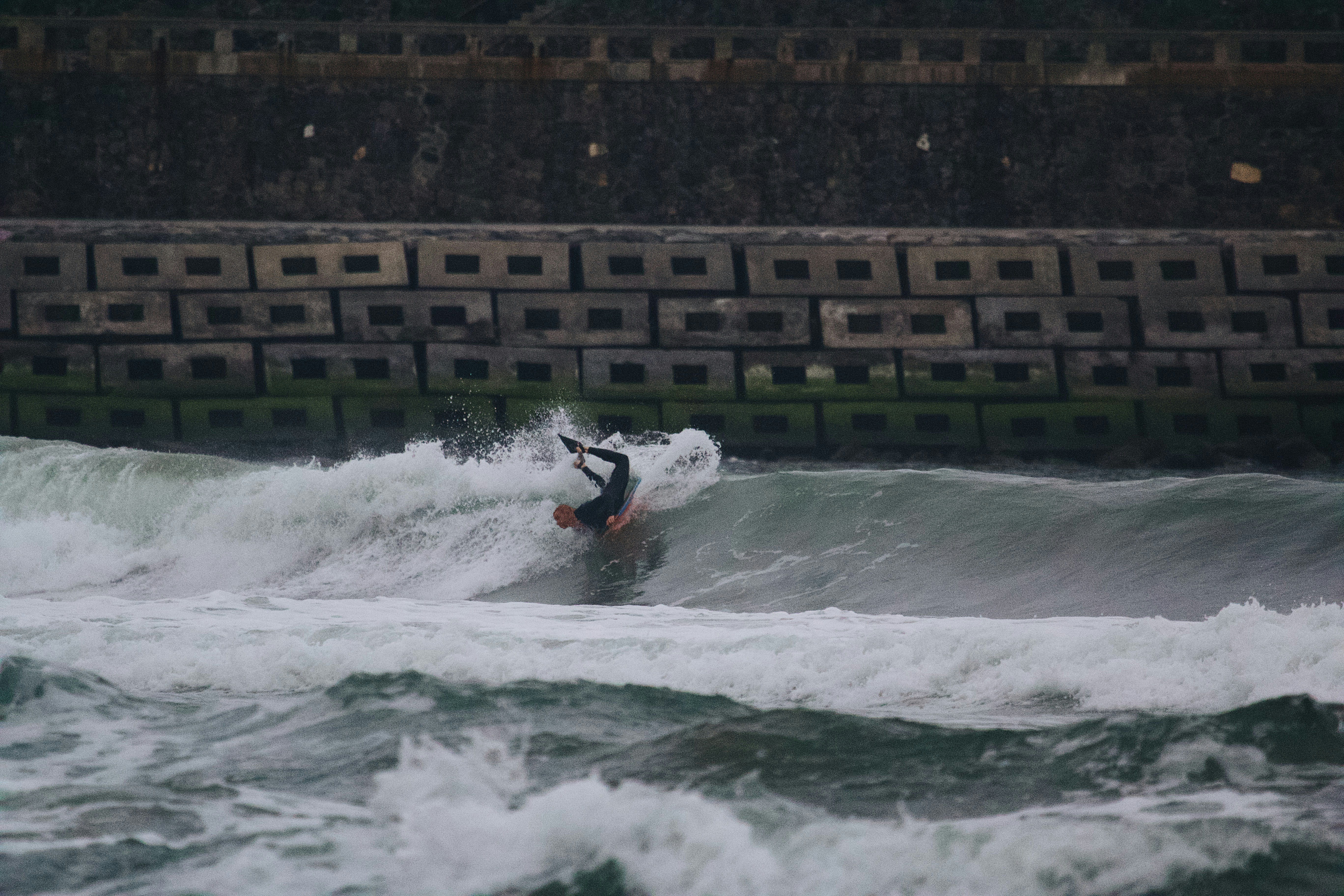 Surfer executing a sharp turn on a wave, with a textured backdrop of a stone pier. The scene captures the energy and movement of the ocean.