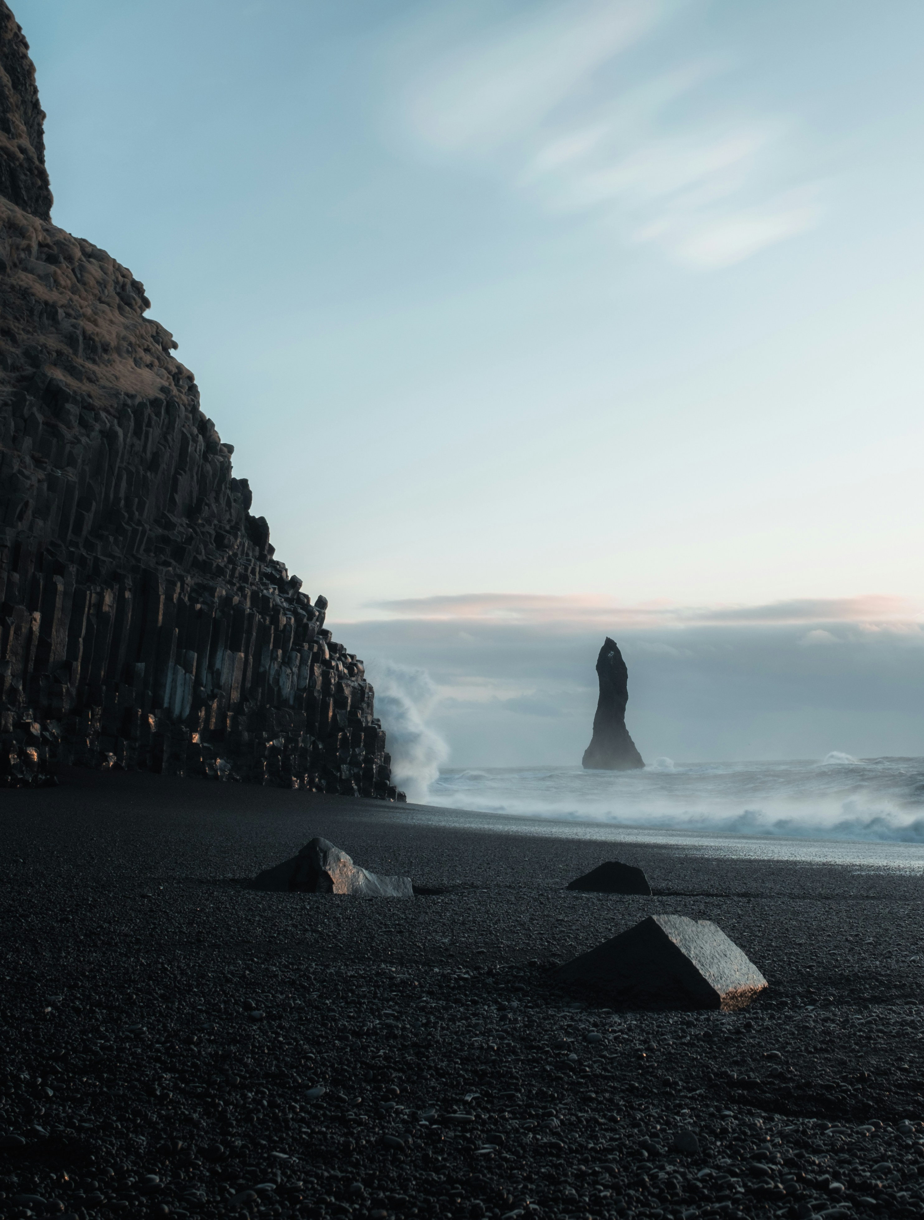 Jagged basalt cliffs rise dramatically beside a tranquil beach, with a solitary sea stack piercing the horizon. Waves gently lap at the shore, creating a serene coastal scene.