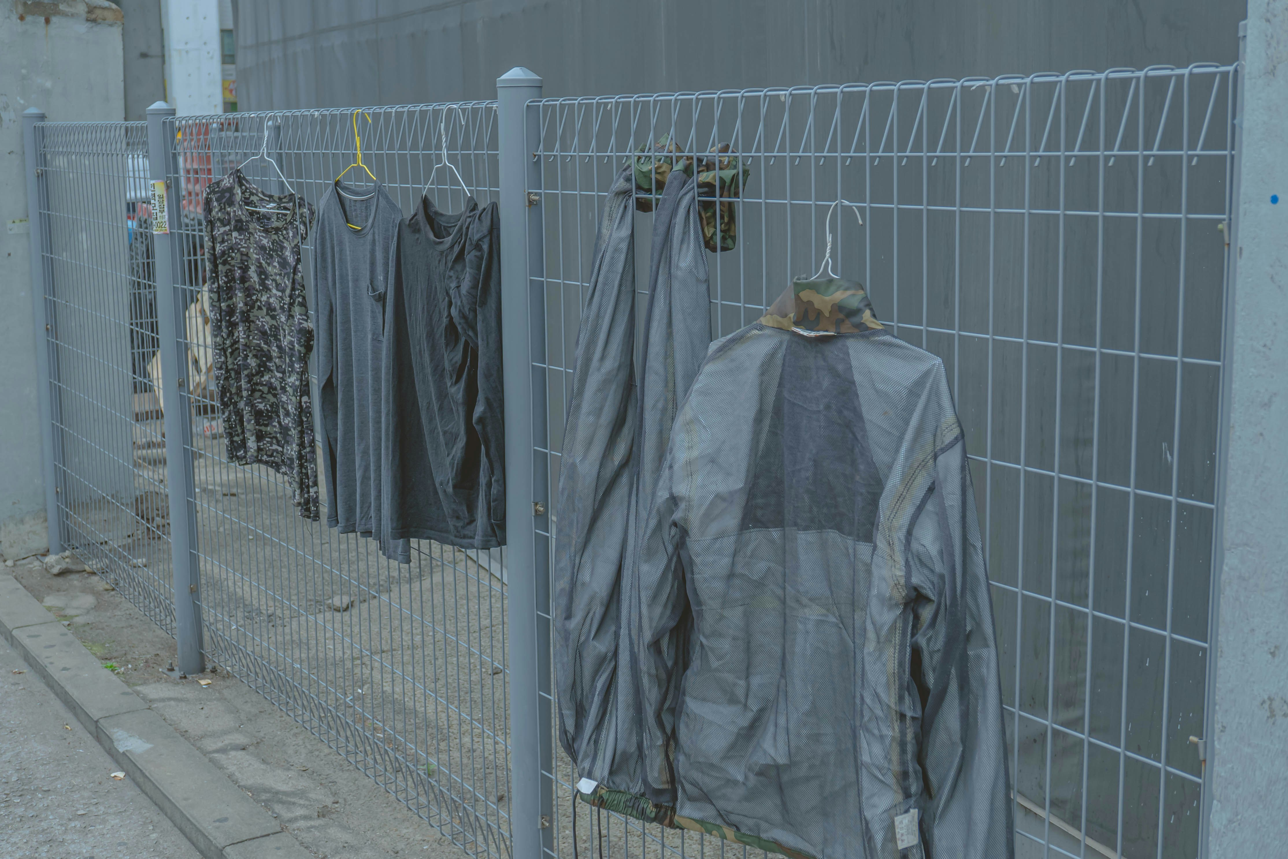 Grey coats and jackets hang along a chain-link fence beside a gray street, creating a quiet, industrial wardrobe scene. The composition emphasizes textures and cool, diffused daylight.