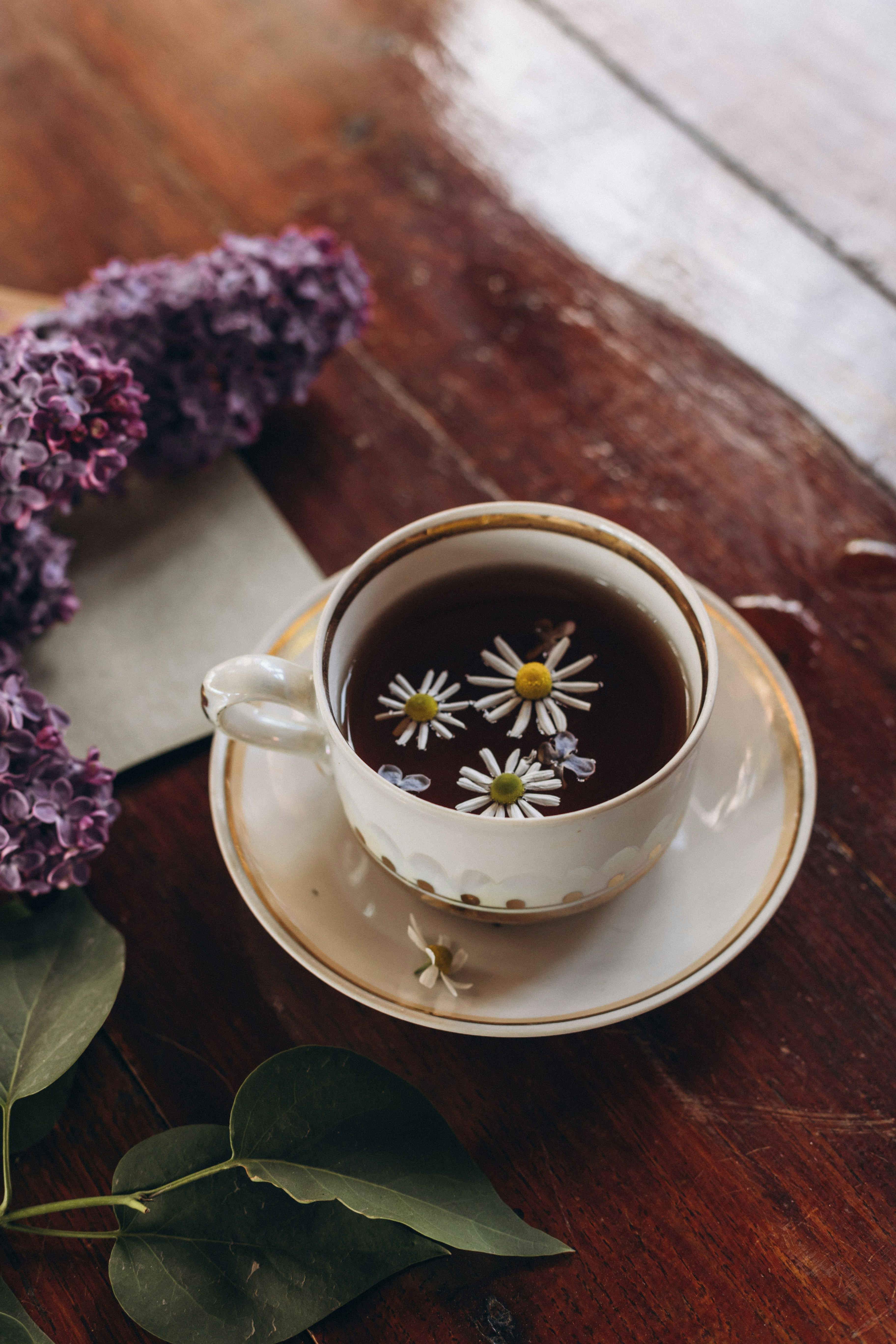 A delicate white cup filled with herbal tea adorned with daisies, accompanied by lilac flowers and green leaves on a wooden table.