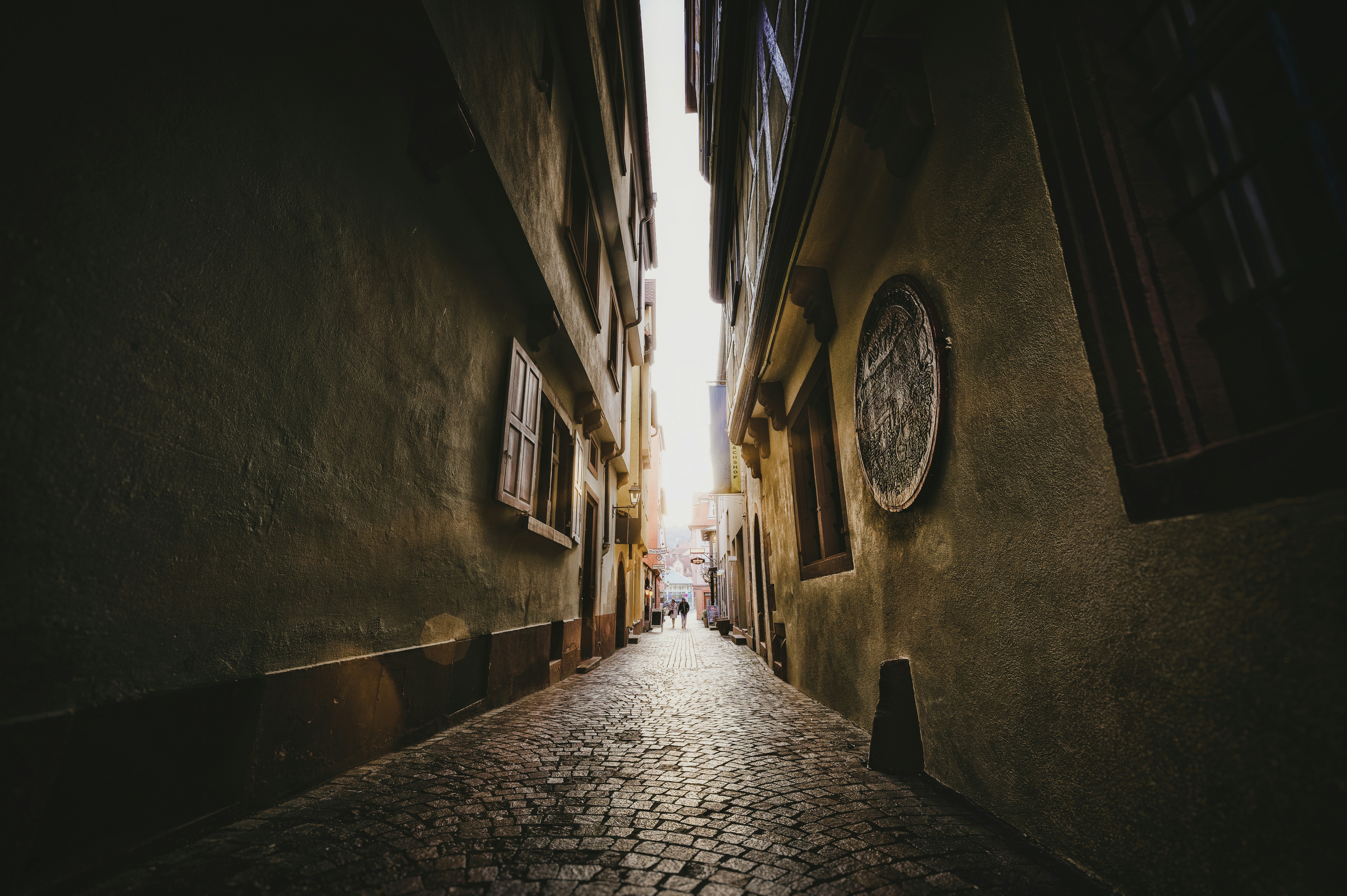 Narrow alley with a wall clock, bordered by tall buildings under soft sunlight.