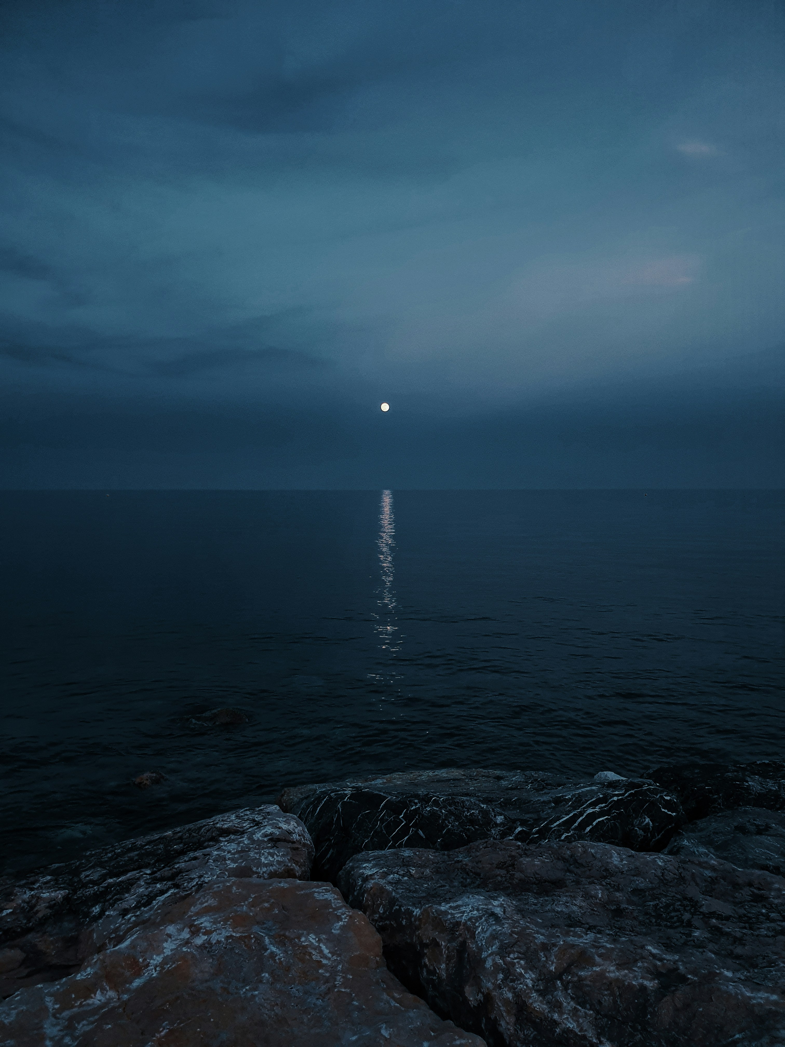 A serene seascape at dusk featuring a bright moon reflecting on calm waters, framed by rocky shoreline.