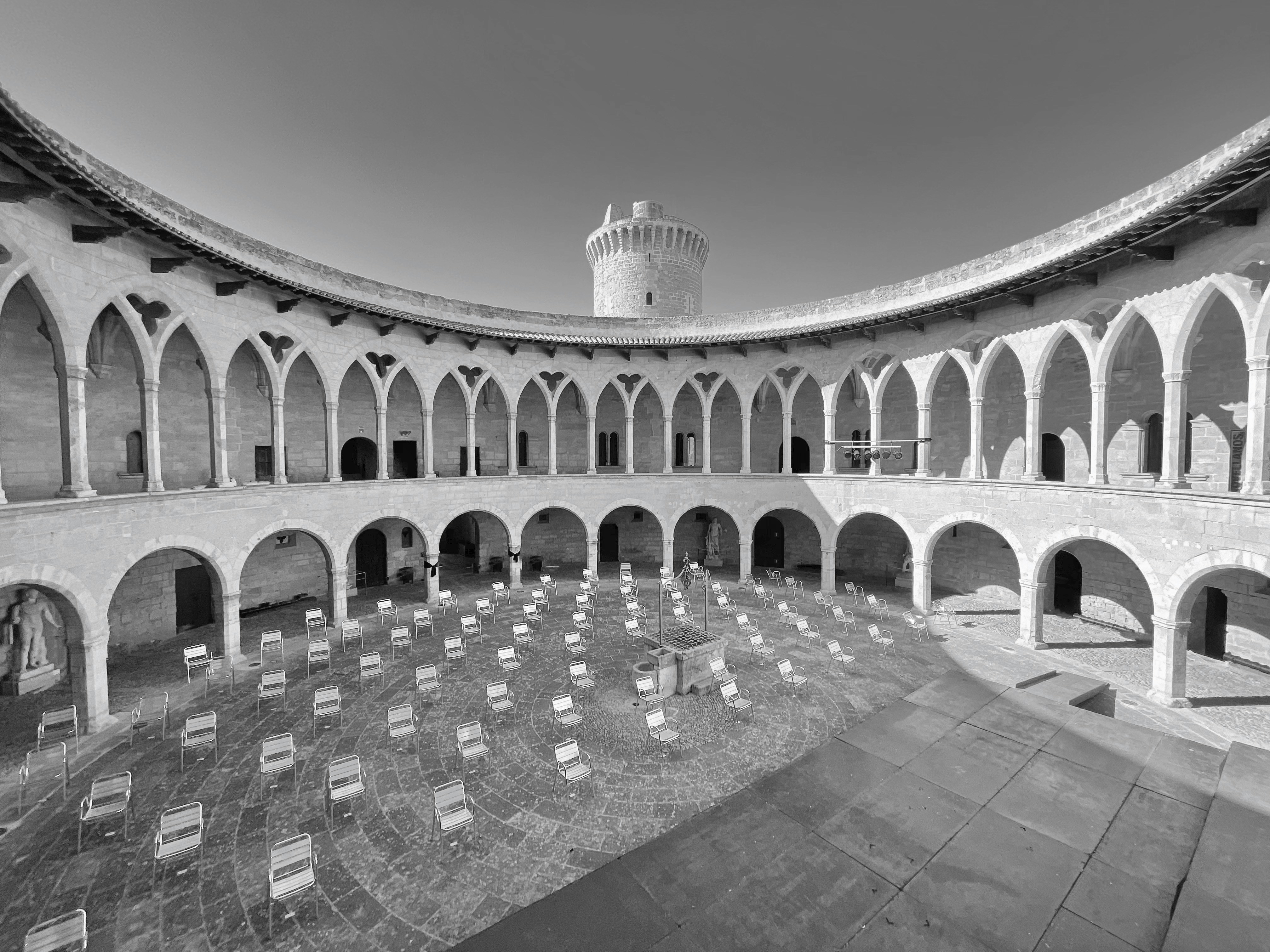 A monochromatic view of a historic courtyard with arched architecture and arranged seating, showcasing a blend of past and present. The tower in the background adds depth to the scene.