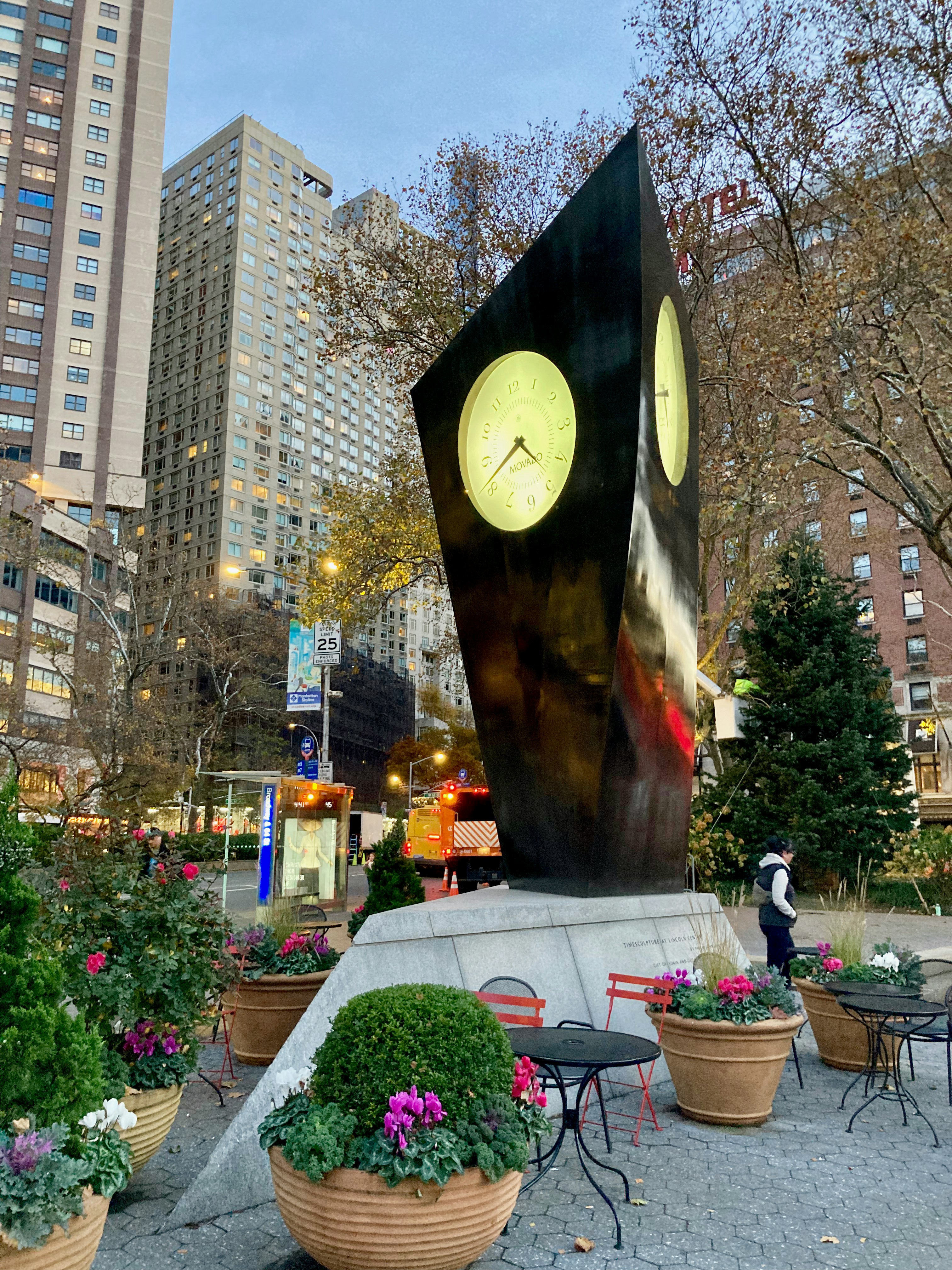 Urban clock sculpture in a city plaza framed by potted greenery