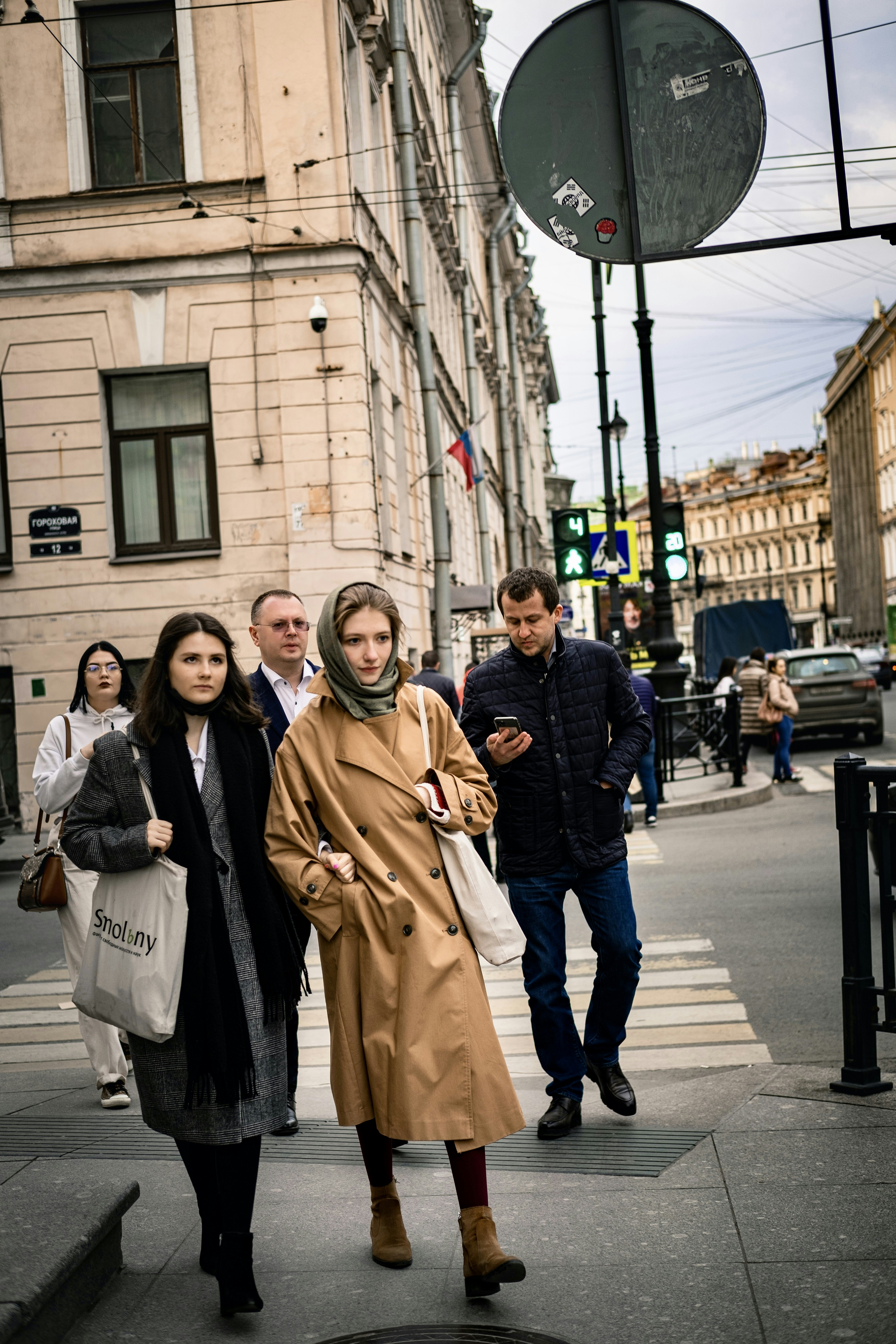 Two women in stylish coats walk confidently across a city street, surrounded by pedestrians and urban architecture. A traffic signal and street signs hint at the bustling environment.